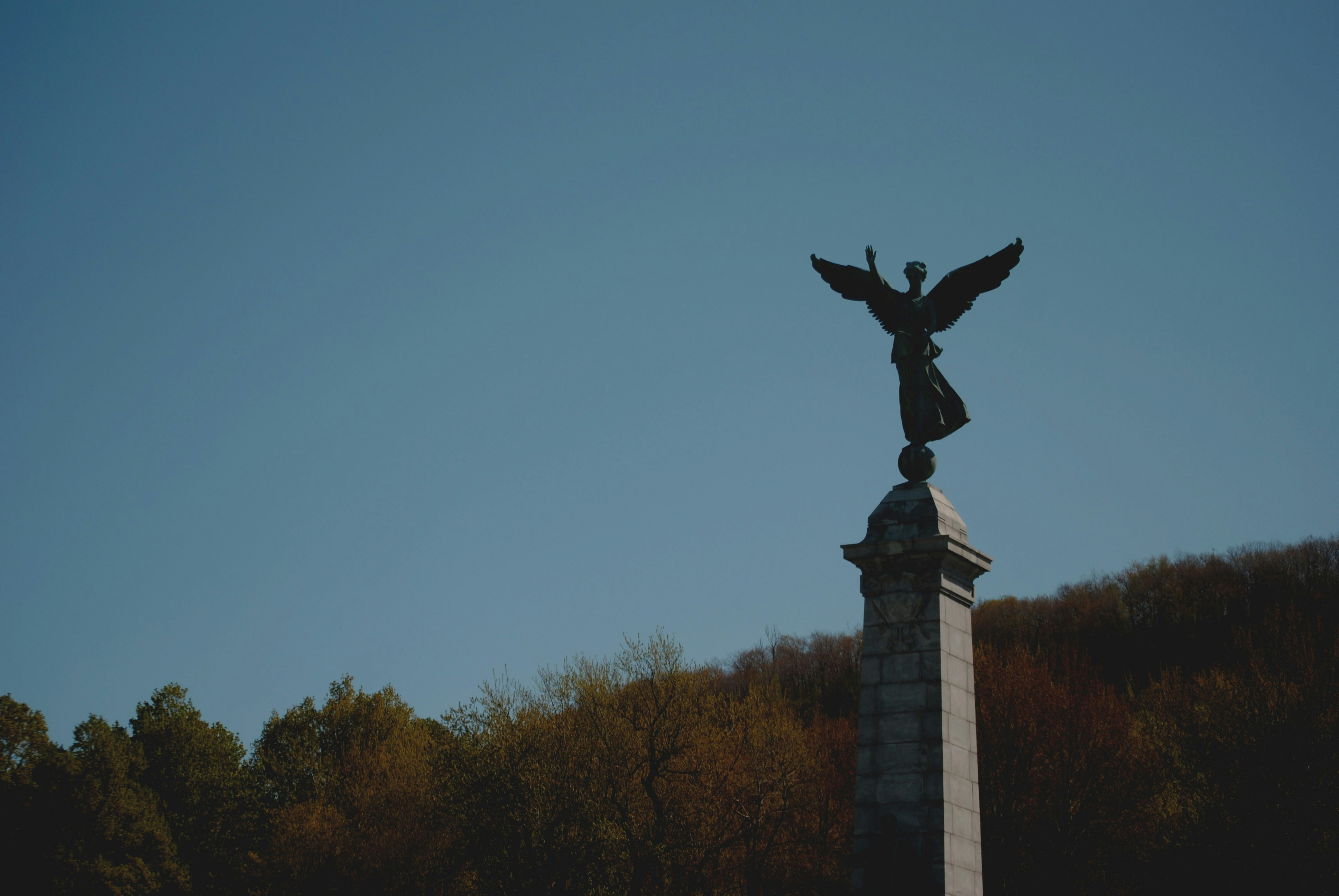 Silhouette of an angel statue atop a stone column against a clear blue sky, surrounded by autumn foliage.