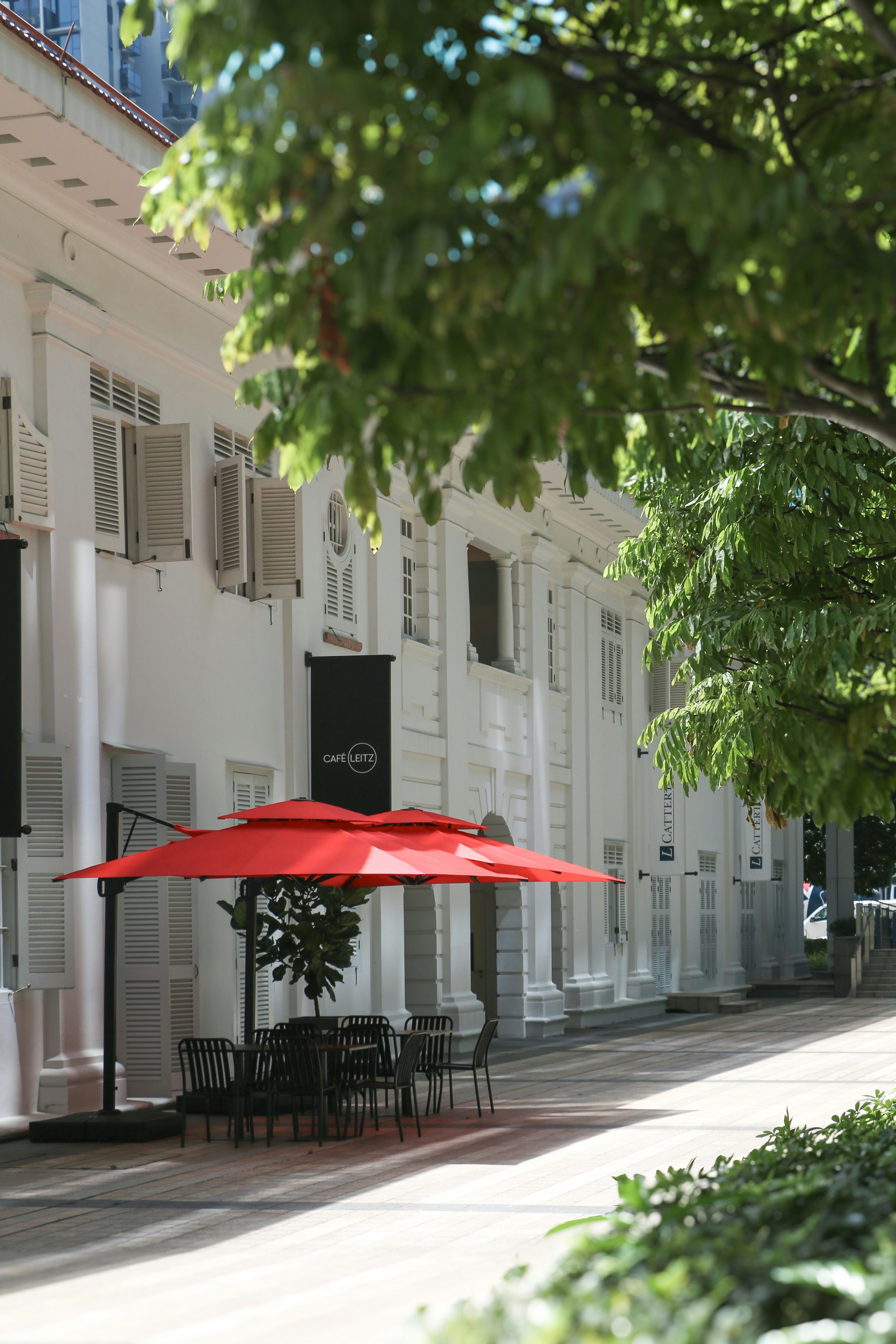 Red umbrella shading a small outdoor café setting against a backdrop of white colonial architecture and lush greenery.