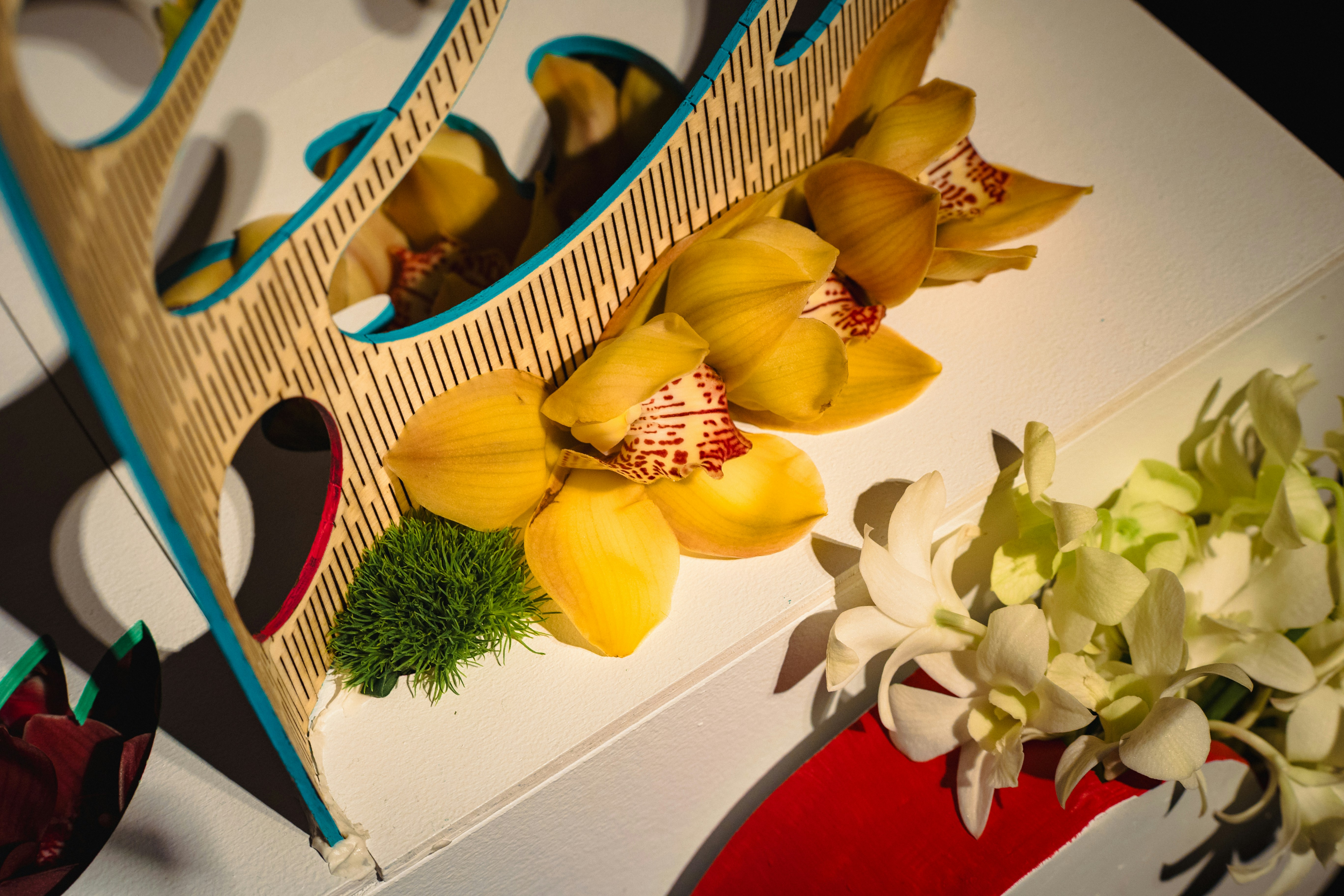 Bright yellow orchids spill from a white and teal decorative basket beside white blossoms on a white surface