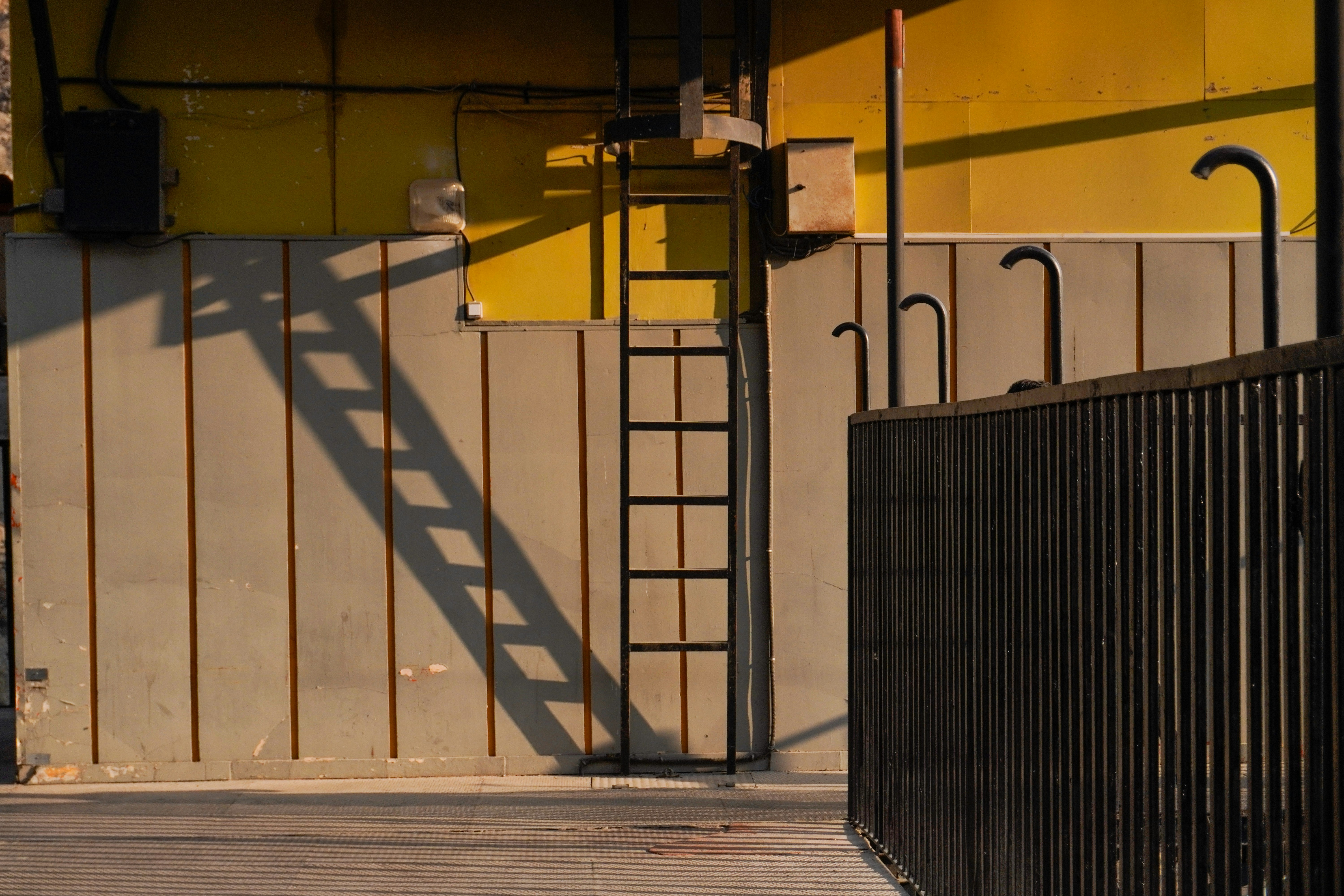 A metal ladder casts a long shadow against a yellow wall, framed by a black railing in an urban setting.