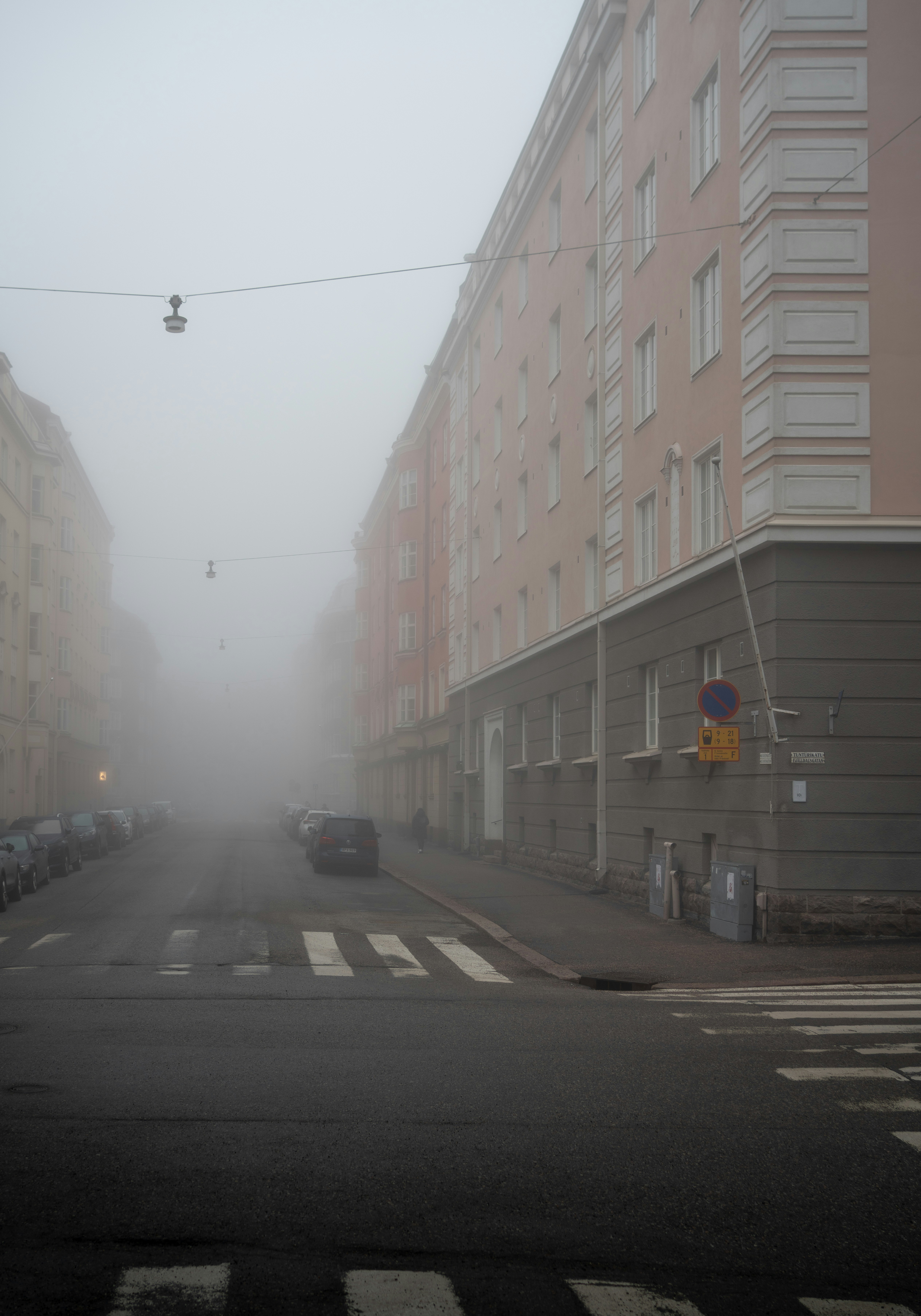 Fog envelops a quiet city street, obscuring the view of parked cars and buildings. The muted colors create an atmospheric scene.