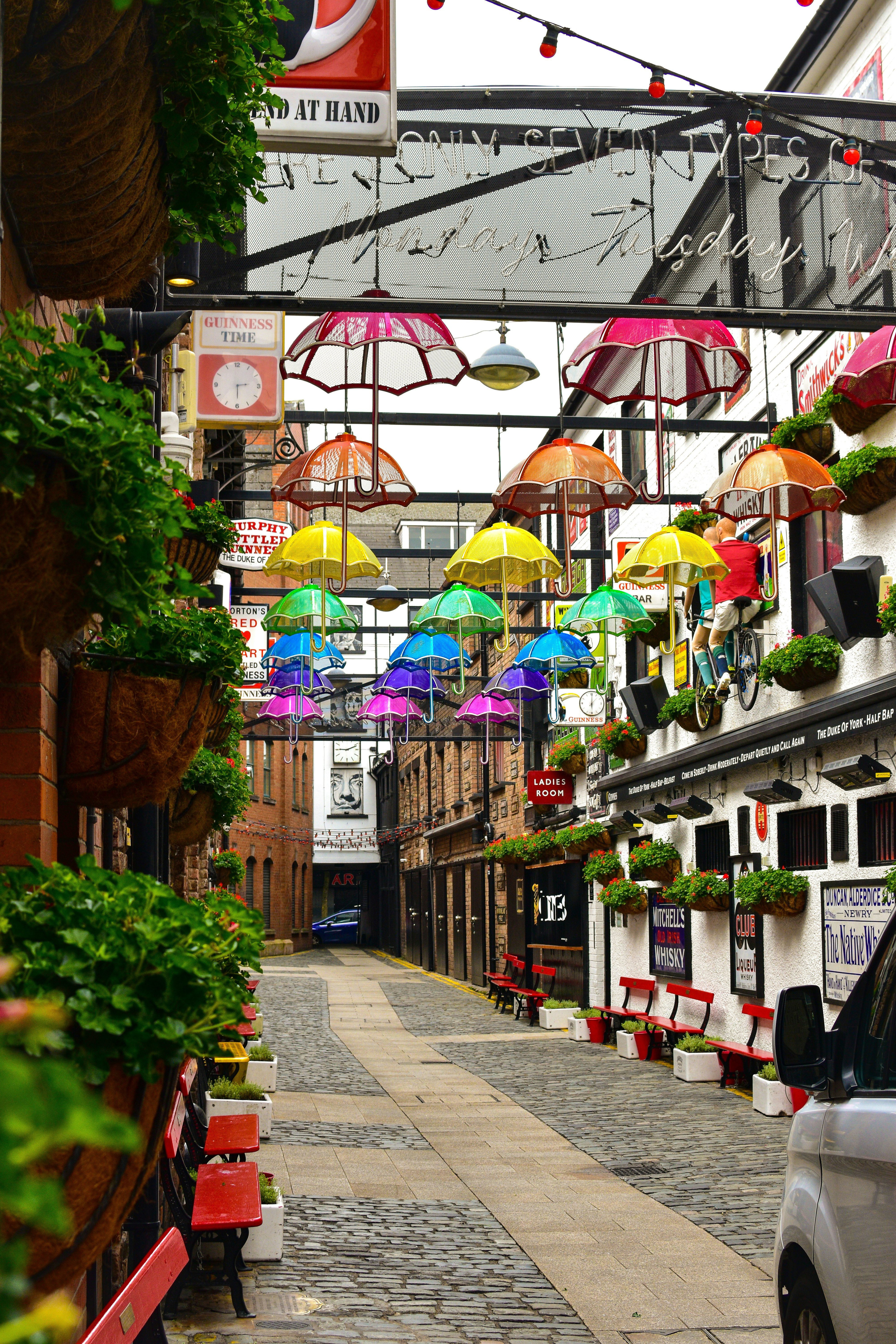 Vibrant umbrellas hang above a charming cobblestone alley, flanked by greenery and rustic signage. The scene captures the lively spirit of the urban environment.