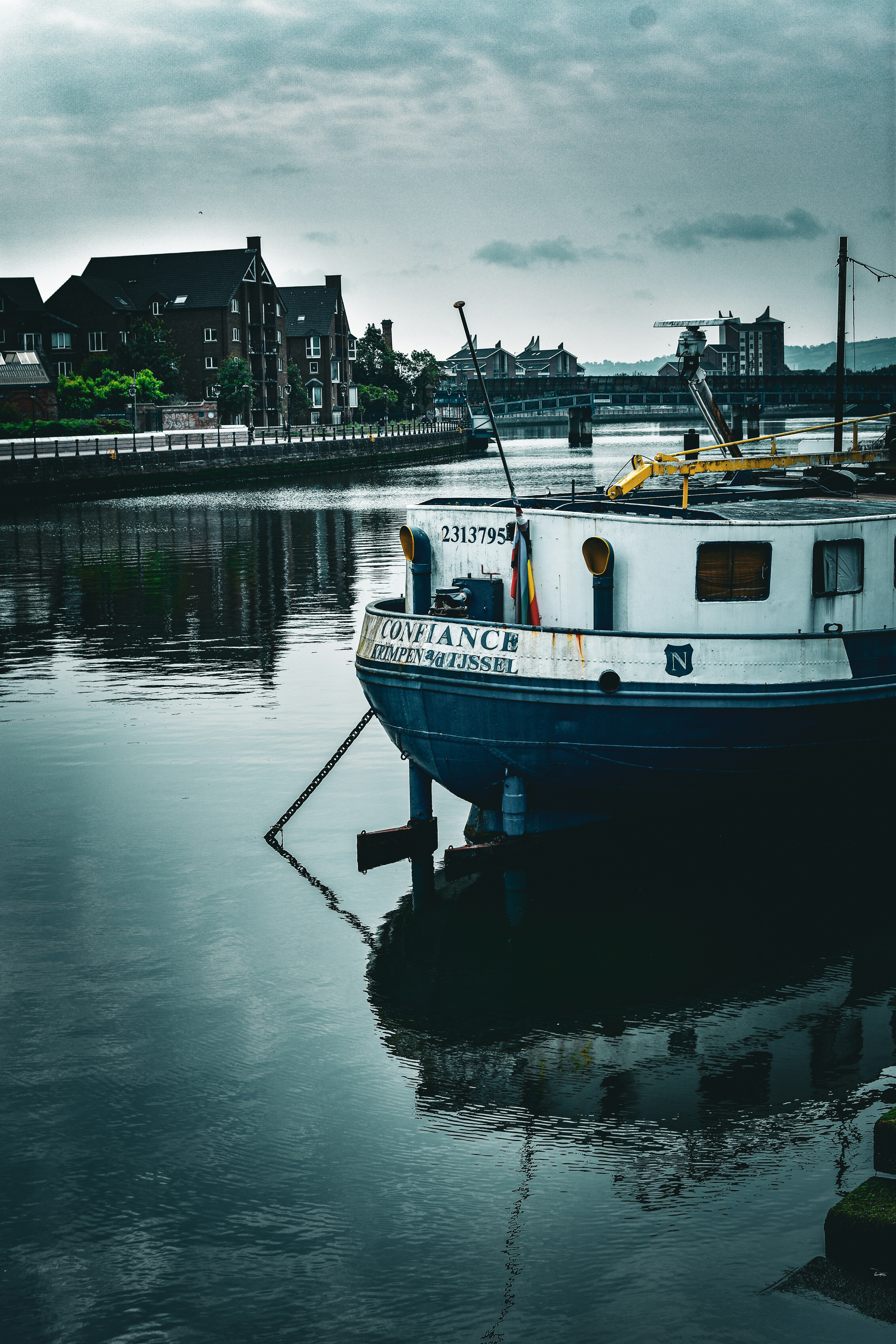 A moored boat named 'Confiance' rests serenely on the still waters, with reflections mirroring the surrounding architecture and cloudy sky.
