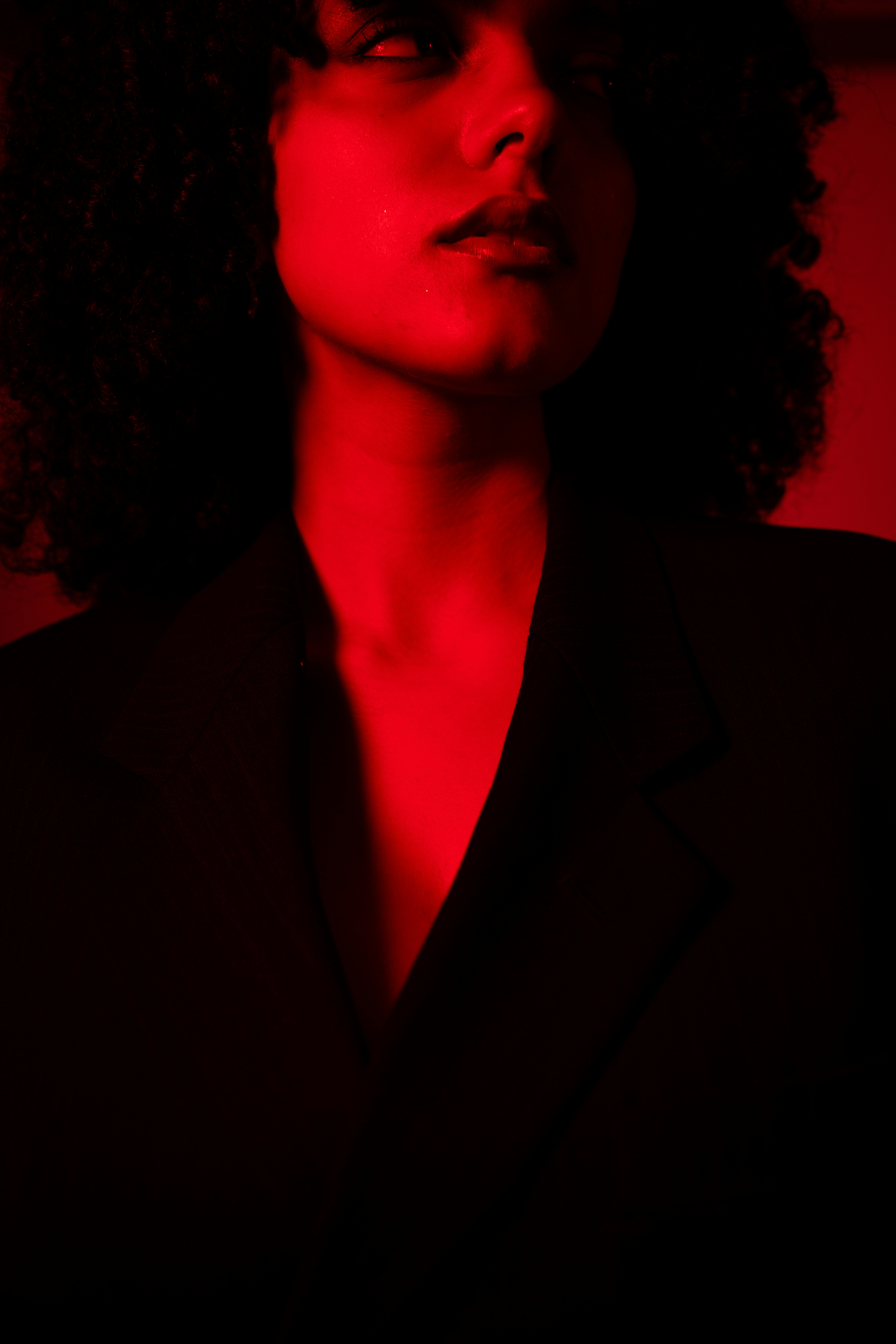A close-up portrait featuring a person with curly hair, illuminated by dramatic red lighting. The subject's expression conveys a sense of intrigue.