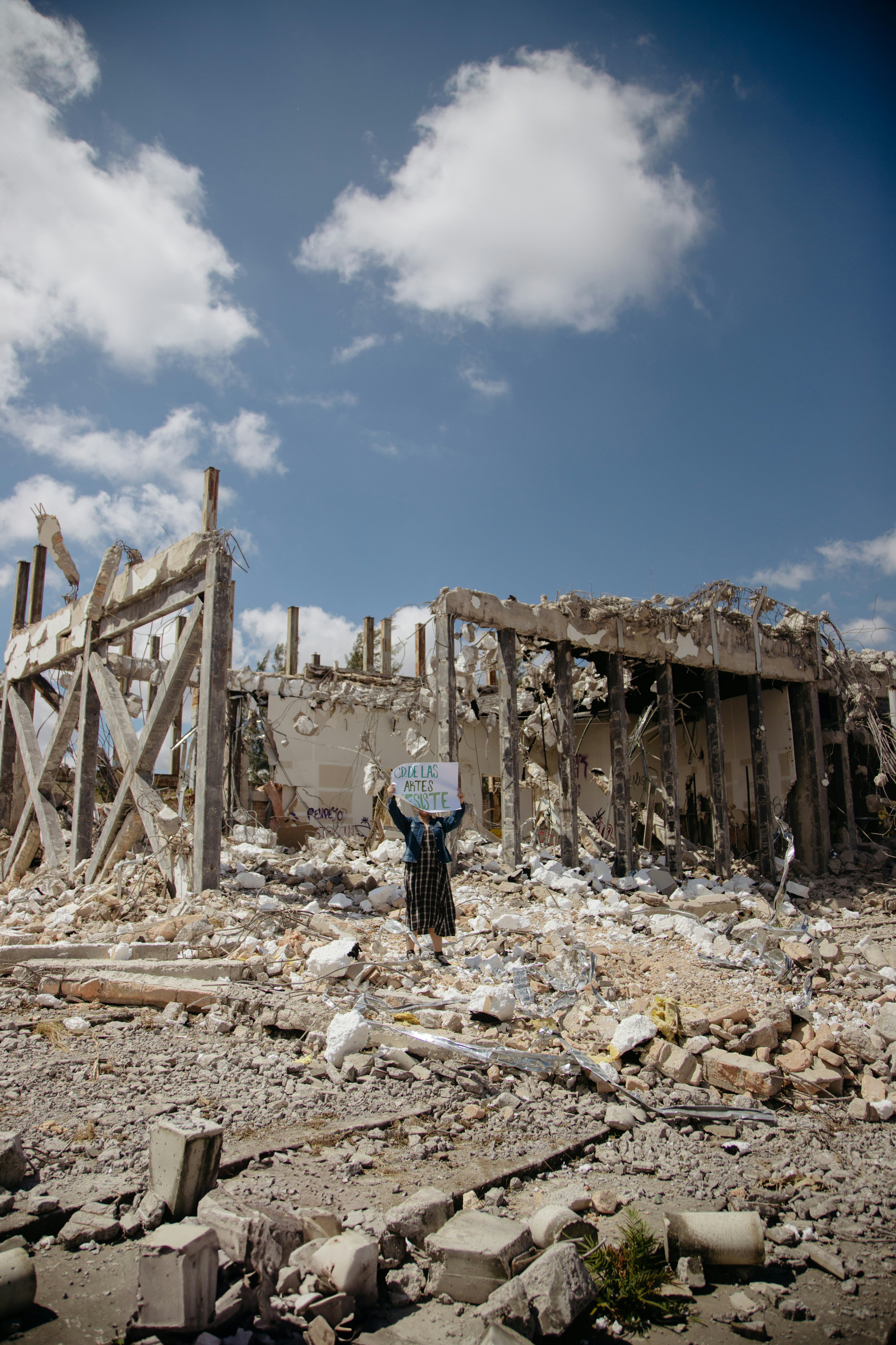 Individual standing in rubble of a collapsed building, holding a sign that conveys a message of hope and resilience.