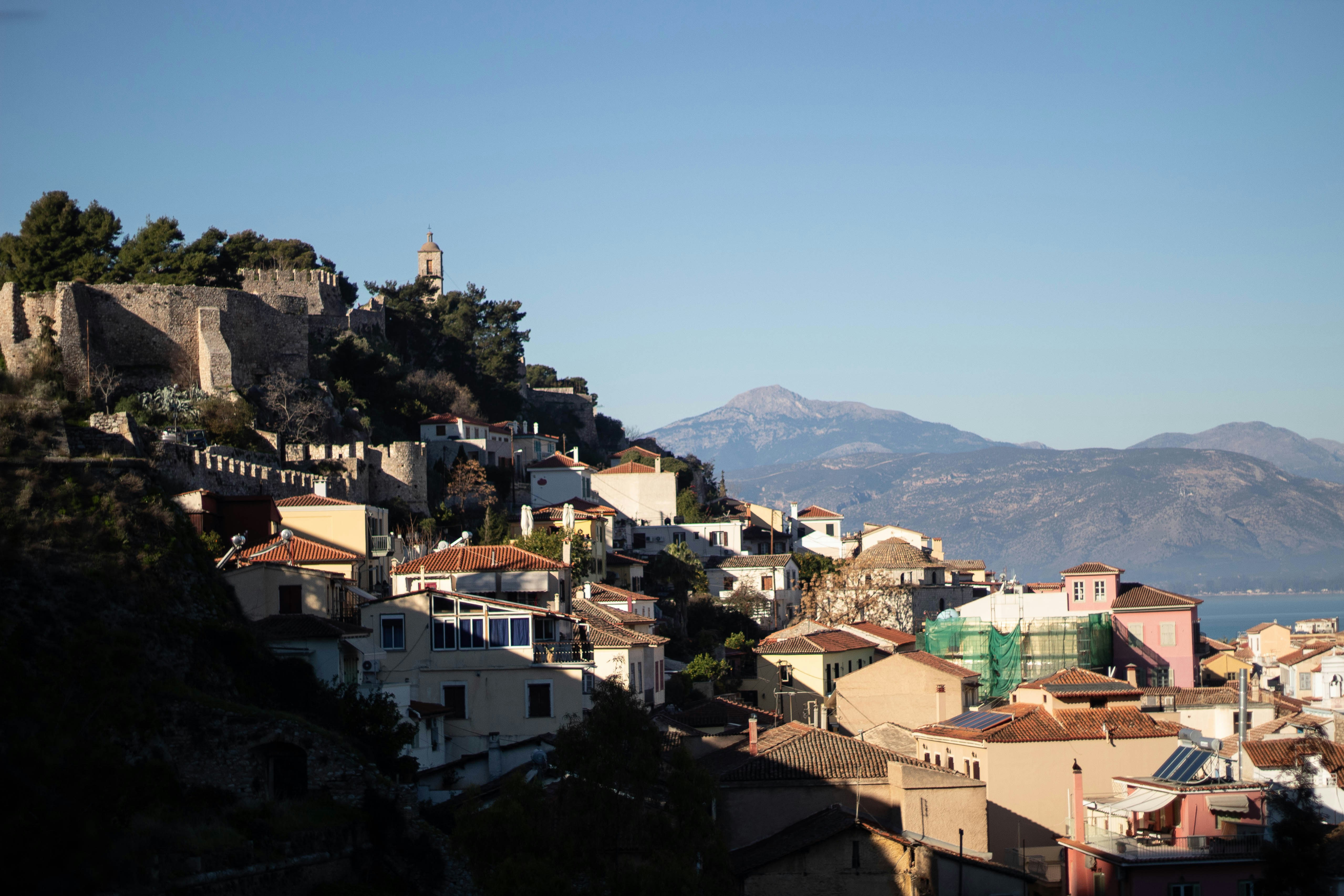 A hillside village featuring terracotta-roofed houses and a historic castle, framed by distant mountains under a clear blue sky.