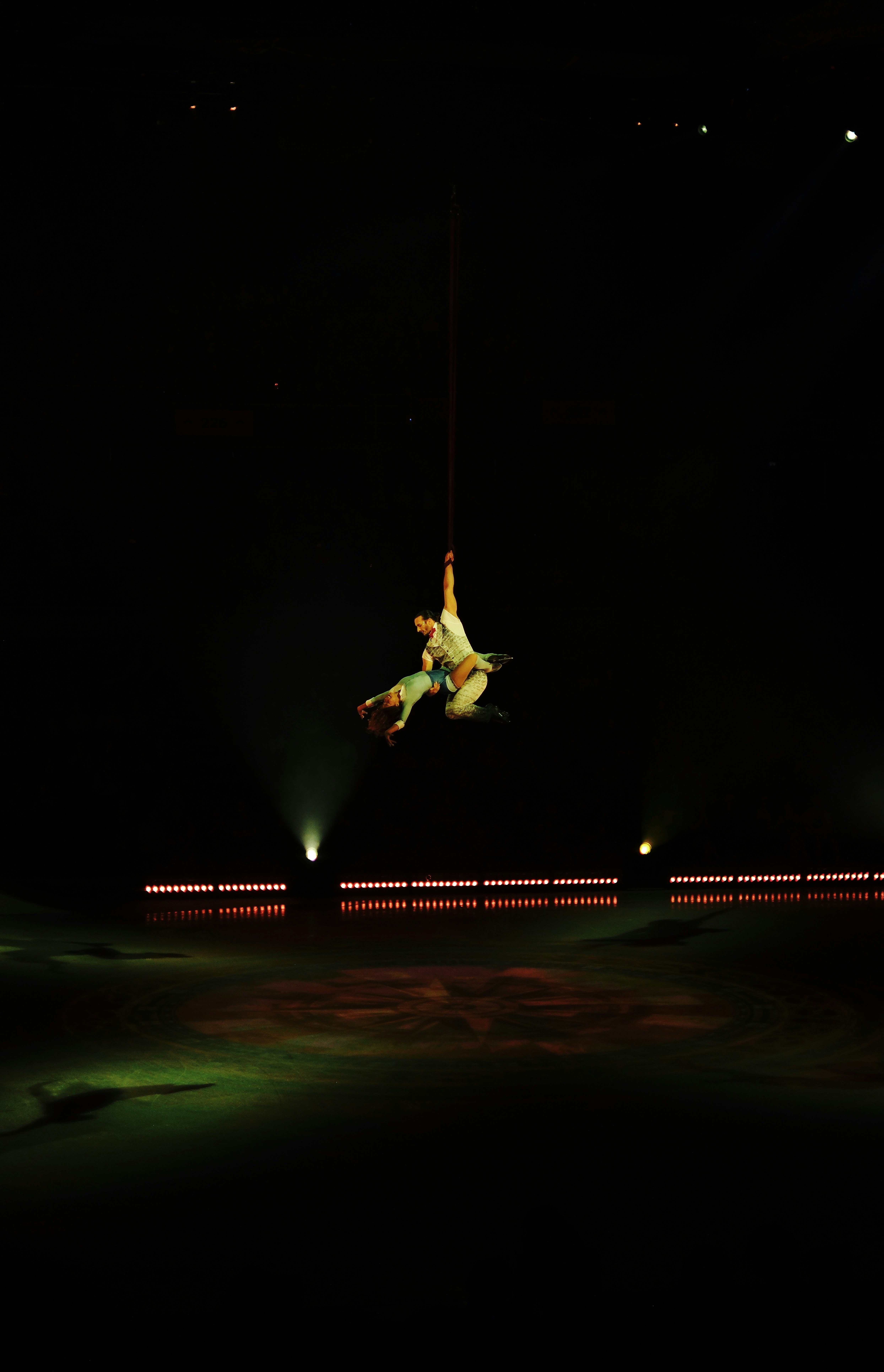 Two aerial performers gracefully suspended from a rope, illuminated by dramatic stage lighting. The scene captures the essence of aerial artistry in motion.