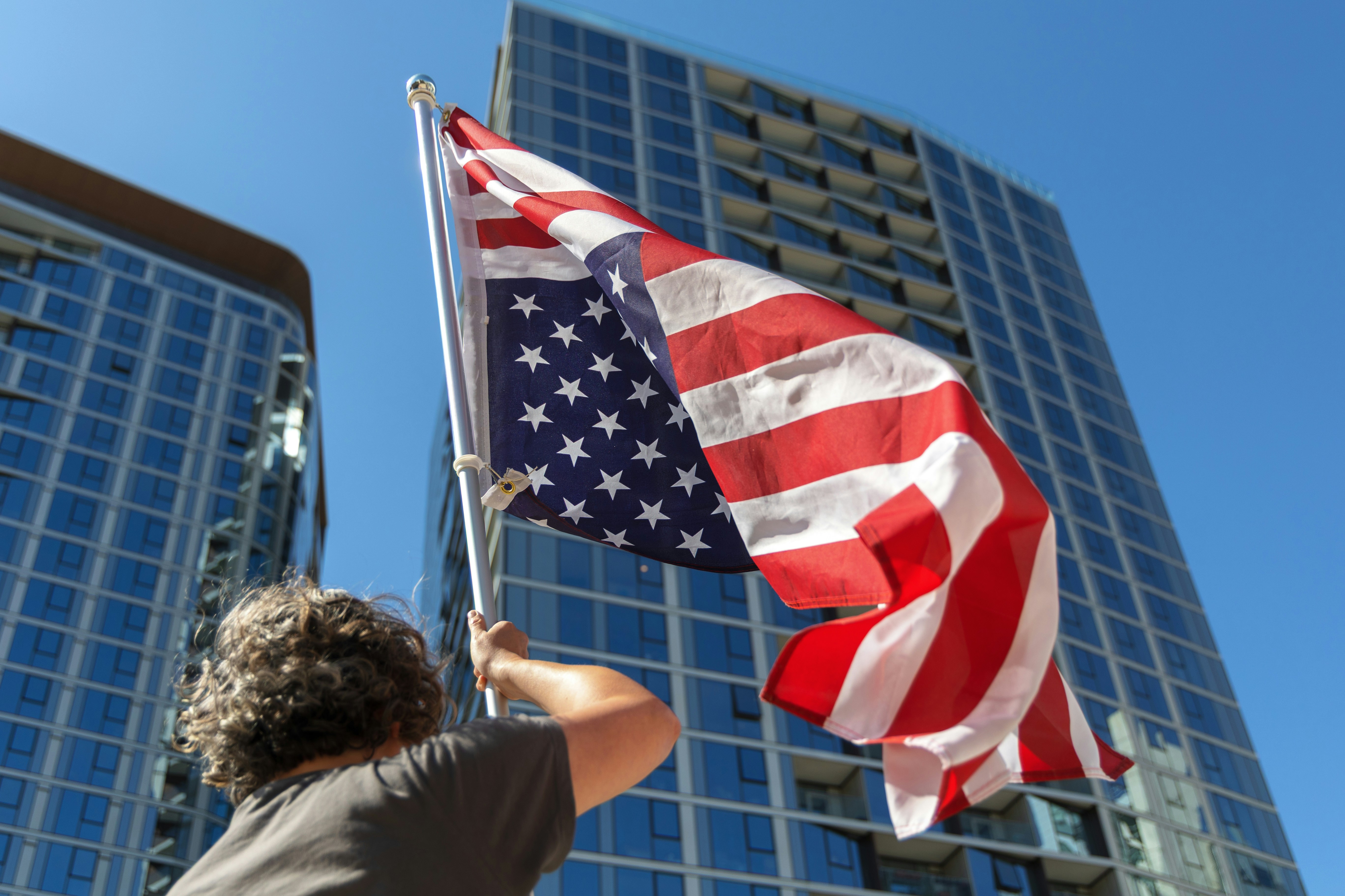 Individual proudly holds an American flag aloft, framed by modern high-rise buildings under a clear blue sky.
