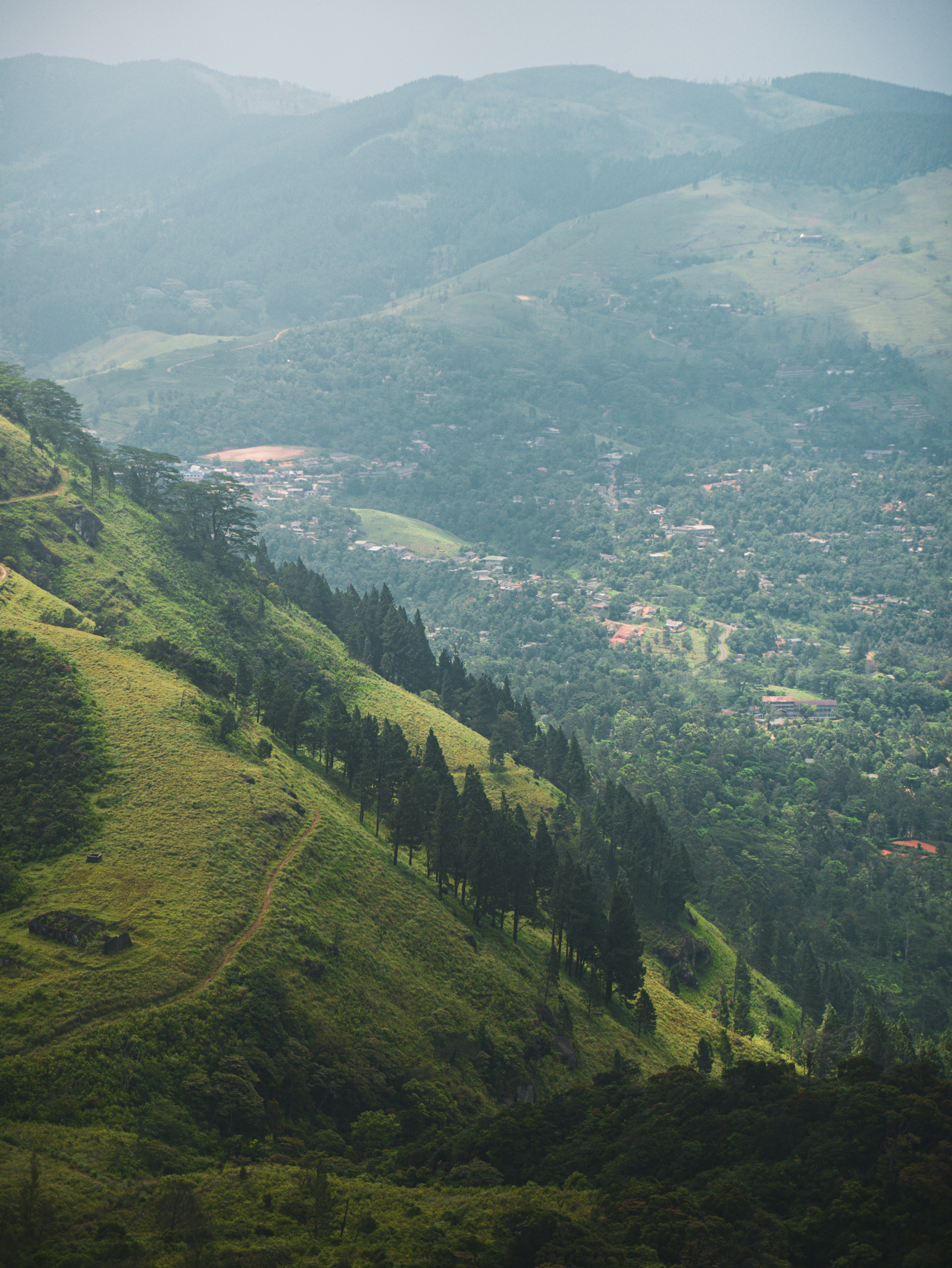 Lush green hills descend into a serene valley, dotted with trees and a glimpse of a small town nestled in the distance.