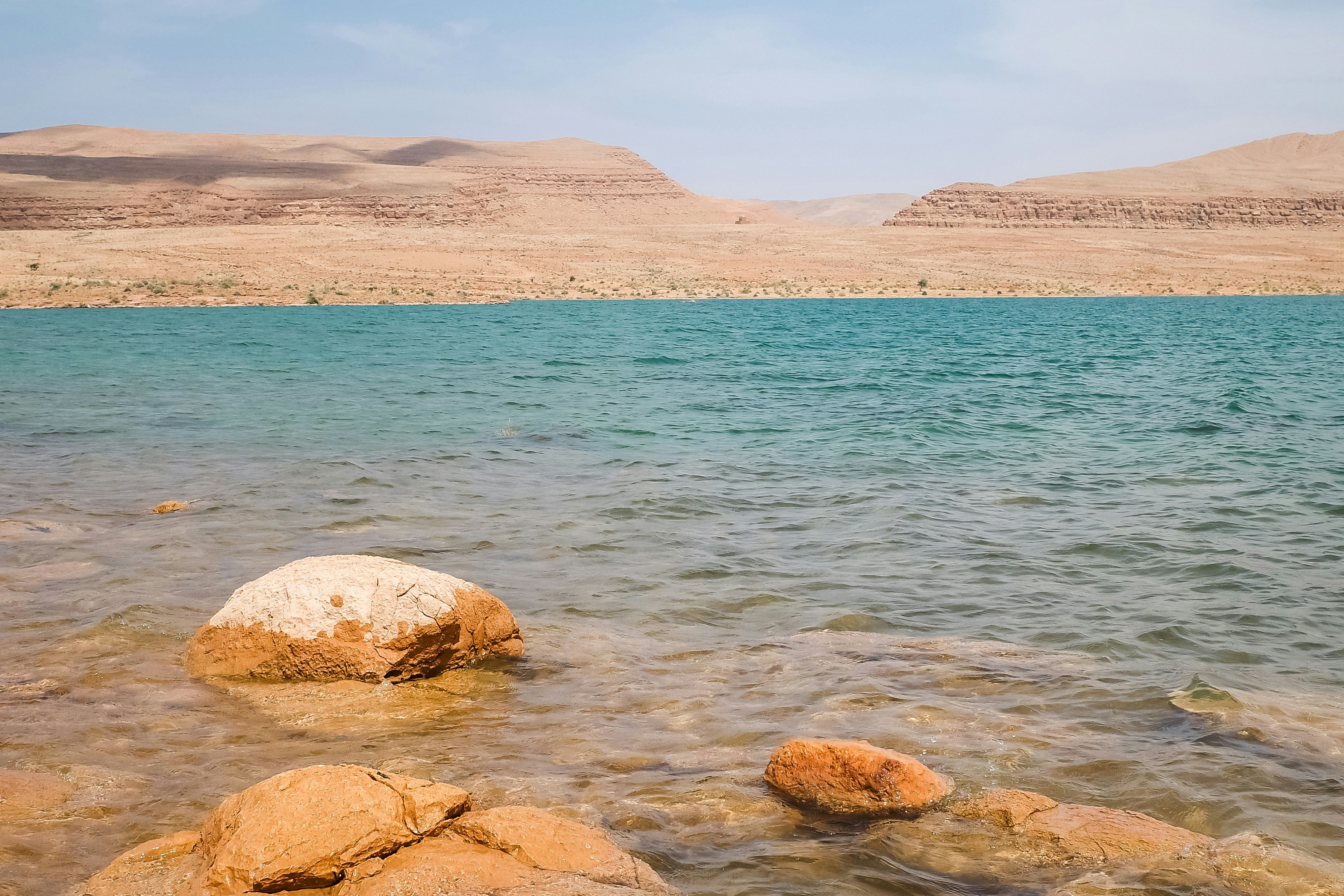 Calm waters reflecting the rugged terrain under a clear sky, with smooth stones lining the shore. A tranquil landscape inviting reflection.