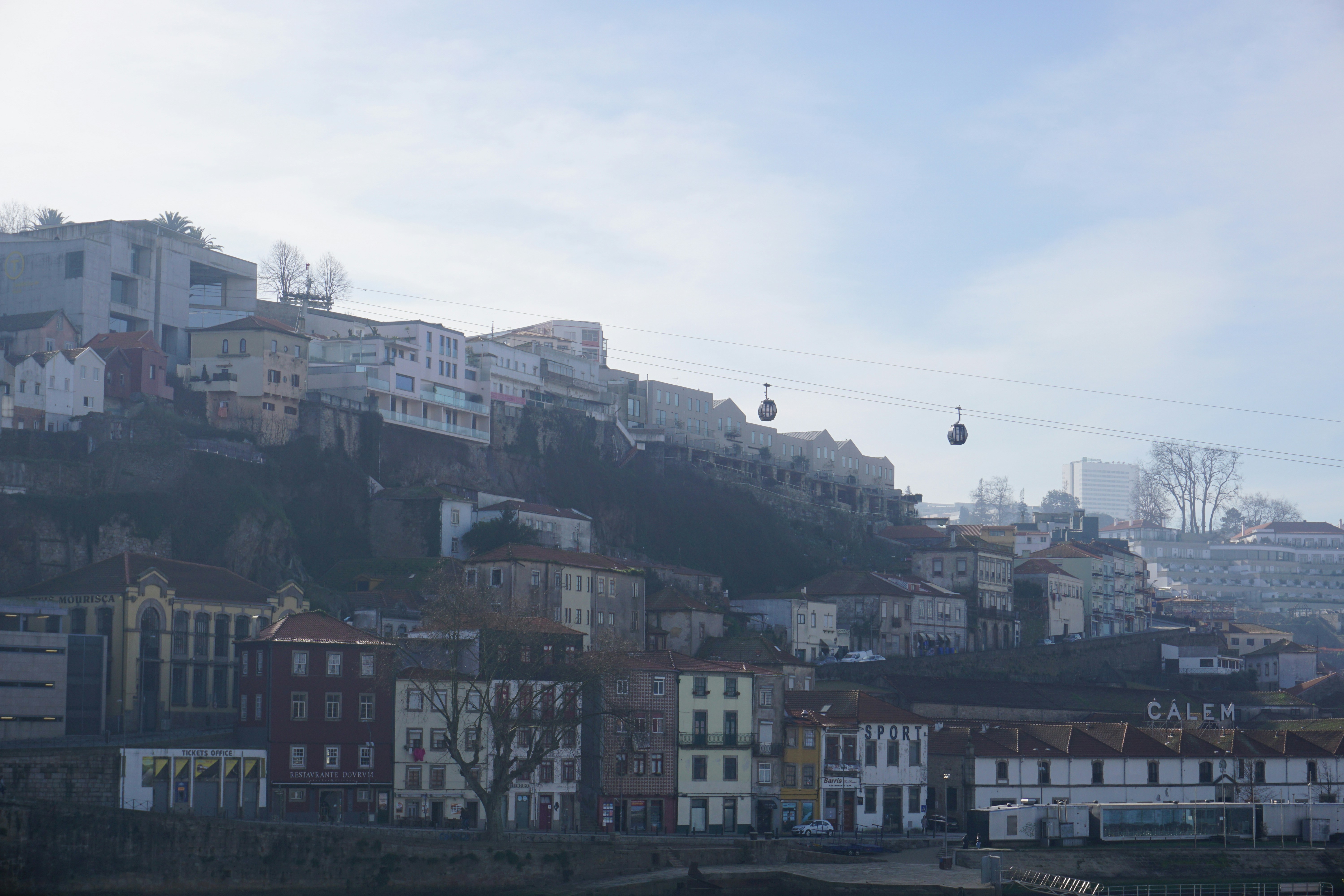 A panoramic view of a hillside adorned with colorful buildings, overlooking a river, with a cable car traversing the skyline.