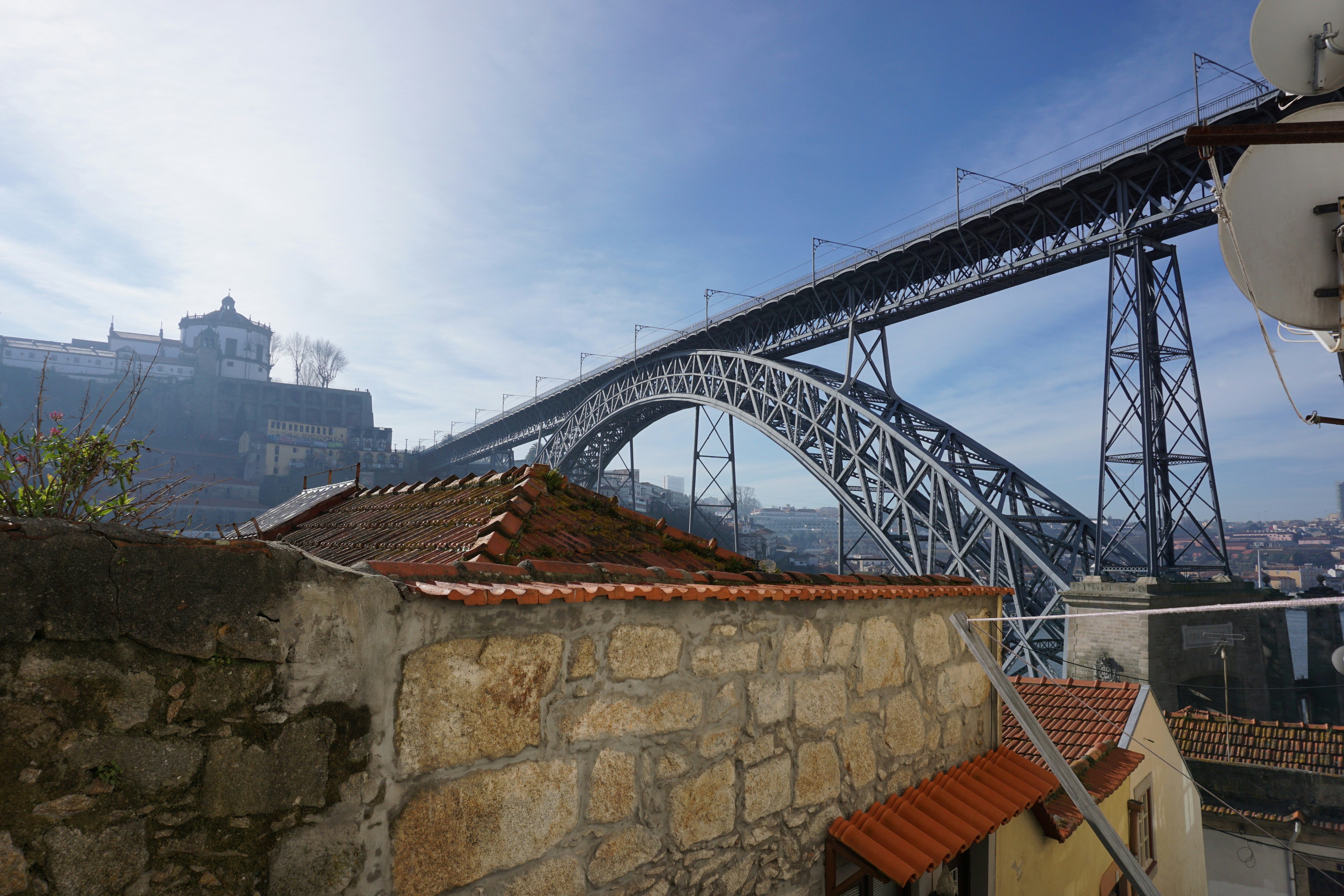 A historic stone wall in the foreground frames the iconic Dom Luís I Bridge spanning the Douro River, with a glimpse of the cityscape beyond.