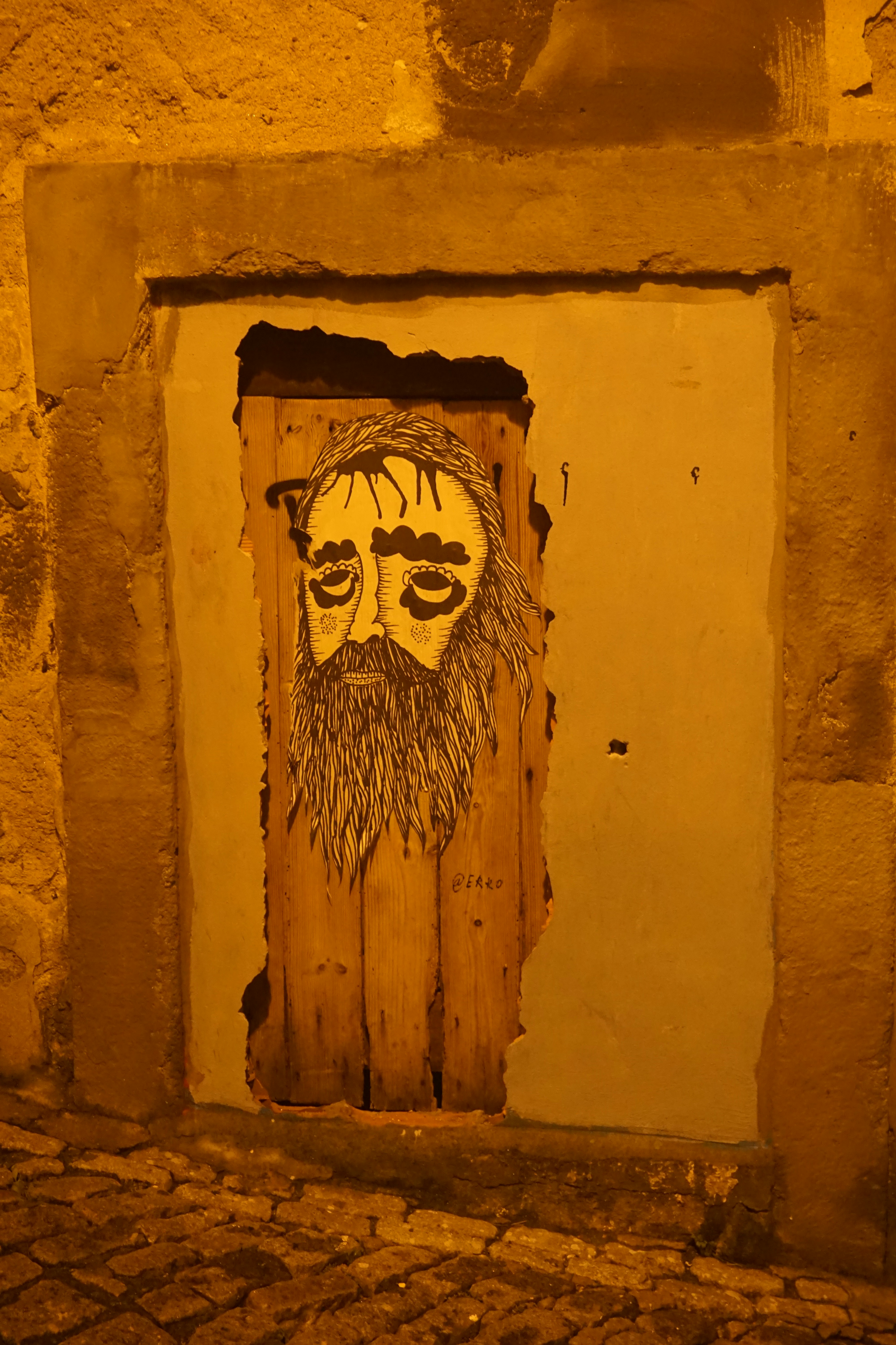 A striking mural of a bearded figure peering through a weathered wooden door, set against a textured wall. The artwork blends street art with urban decay.
