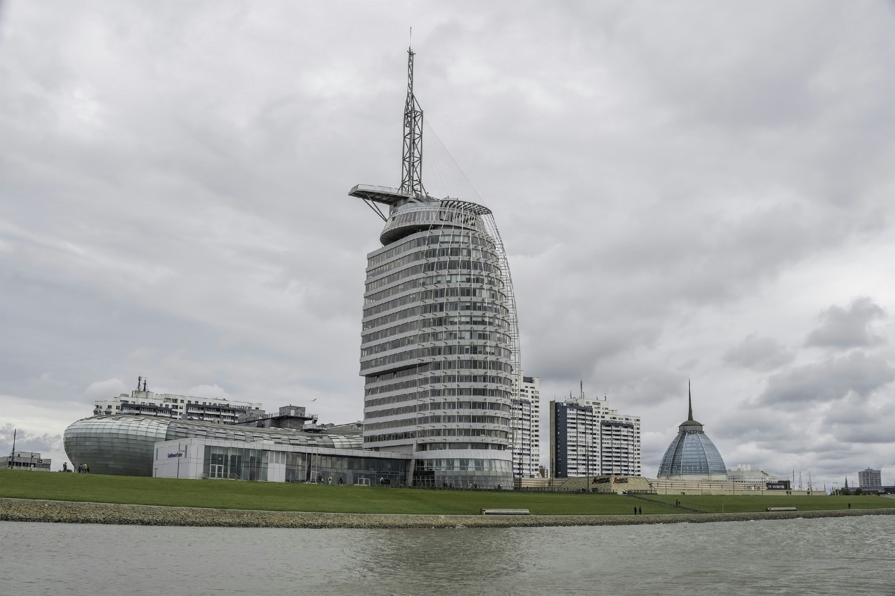 Modern skyscraper with a unique design alongside other contemporary buildings by the river. The cloudy sky adds depth to the urban landscape.