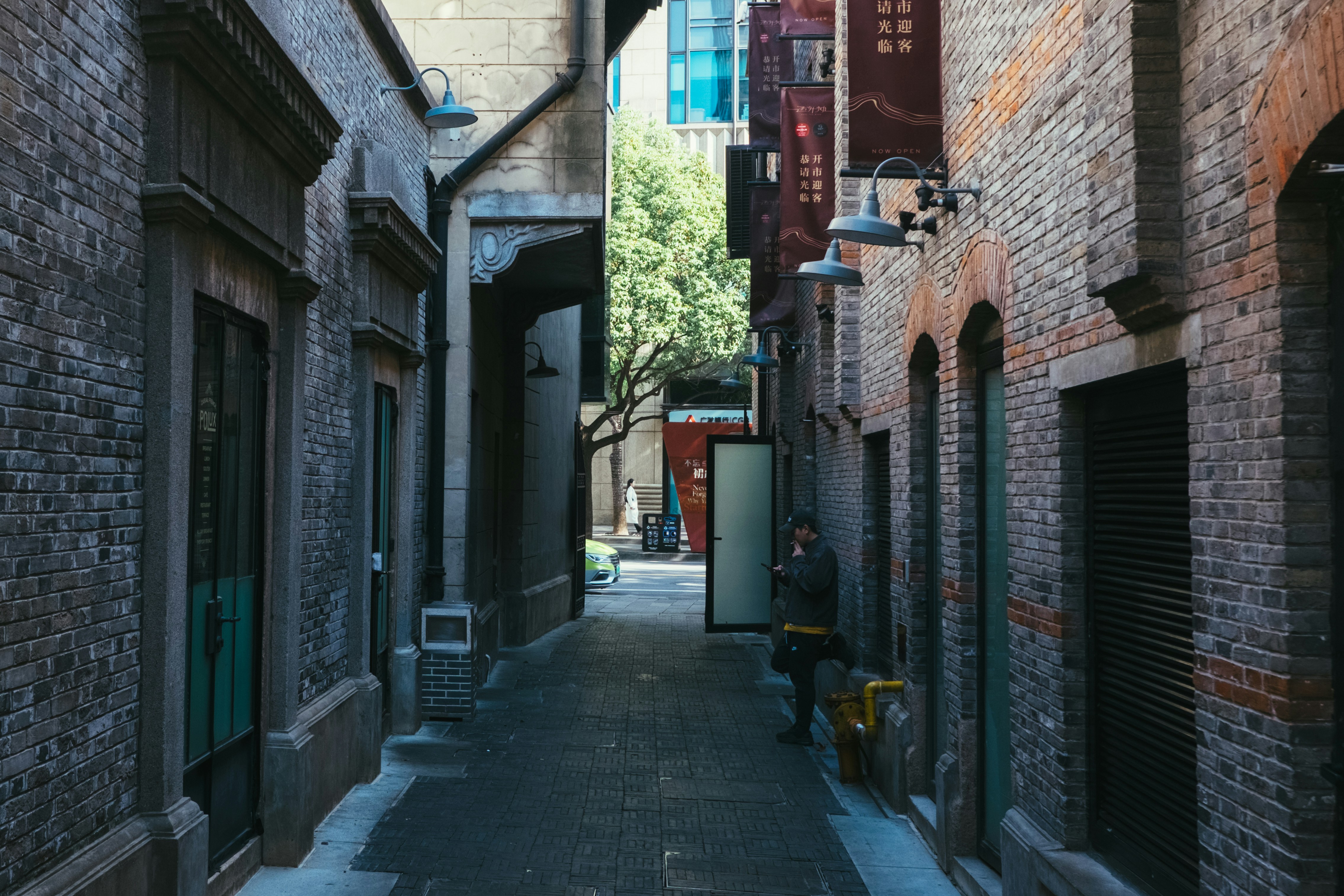 Narrow alleyway flanked by brick buildings, featuring a lone figure absorbed in thought. Lush greenery peeks from the end of the passage.