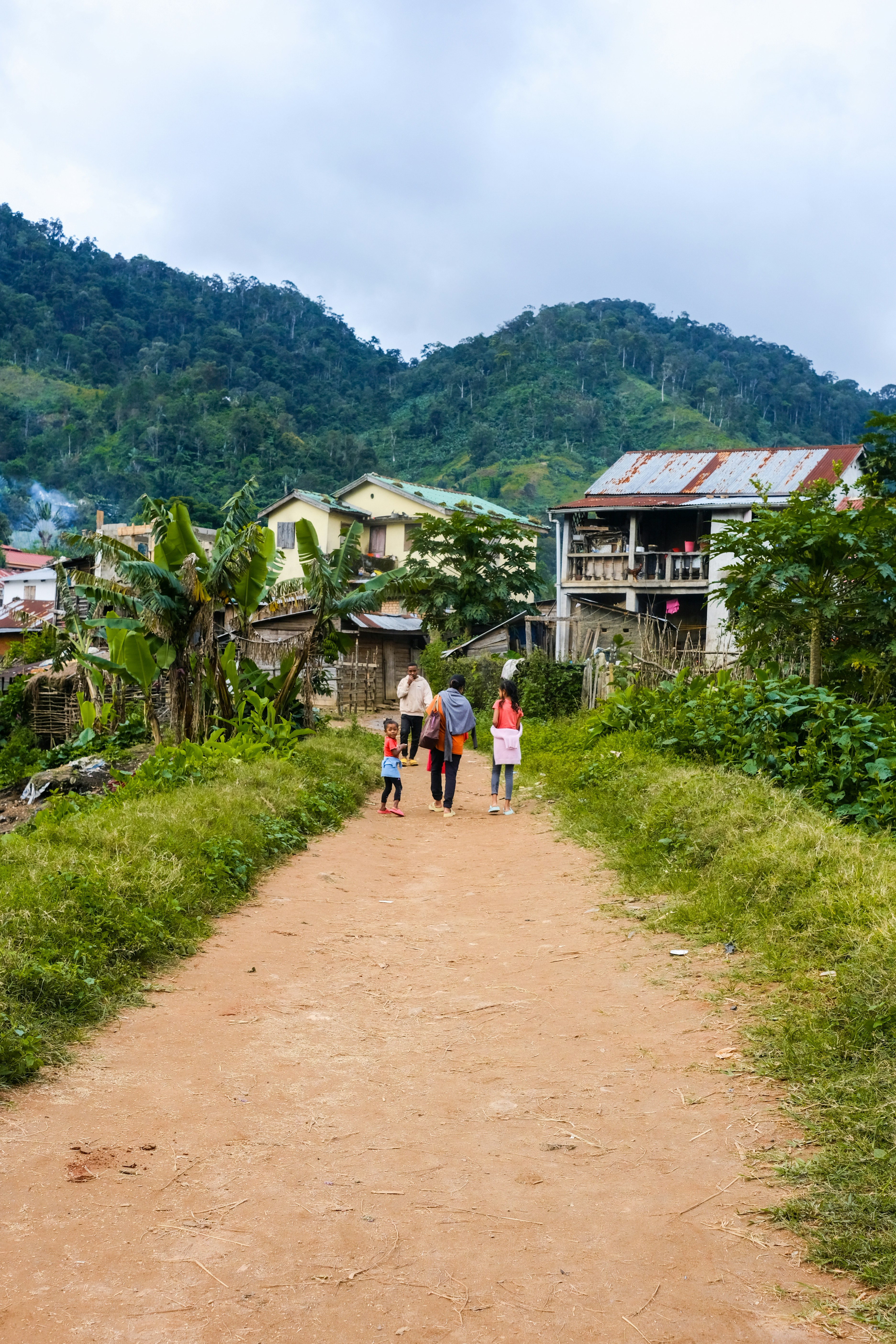 Four individuals walking along a dirt path flanked by lush greenery and rustic homes, set against a backdrop of rolling hills. The scene captures the essence of rural life.