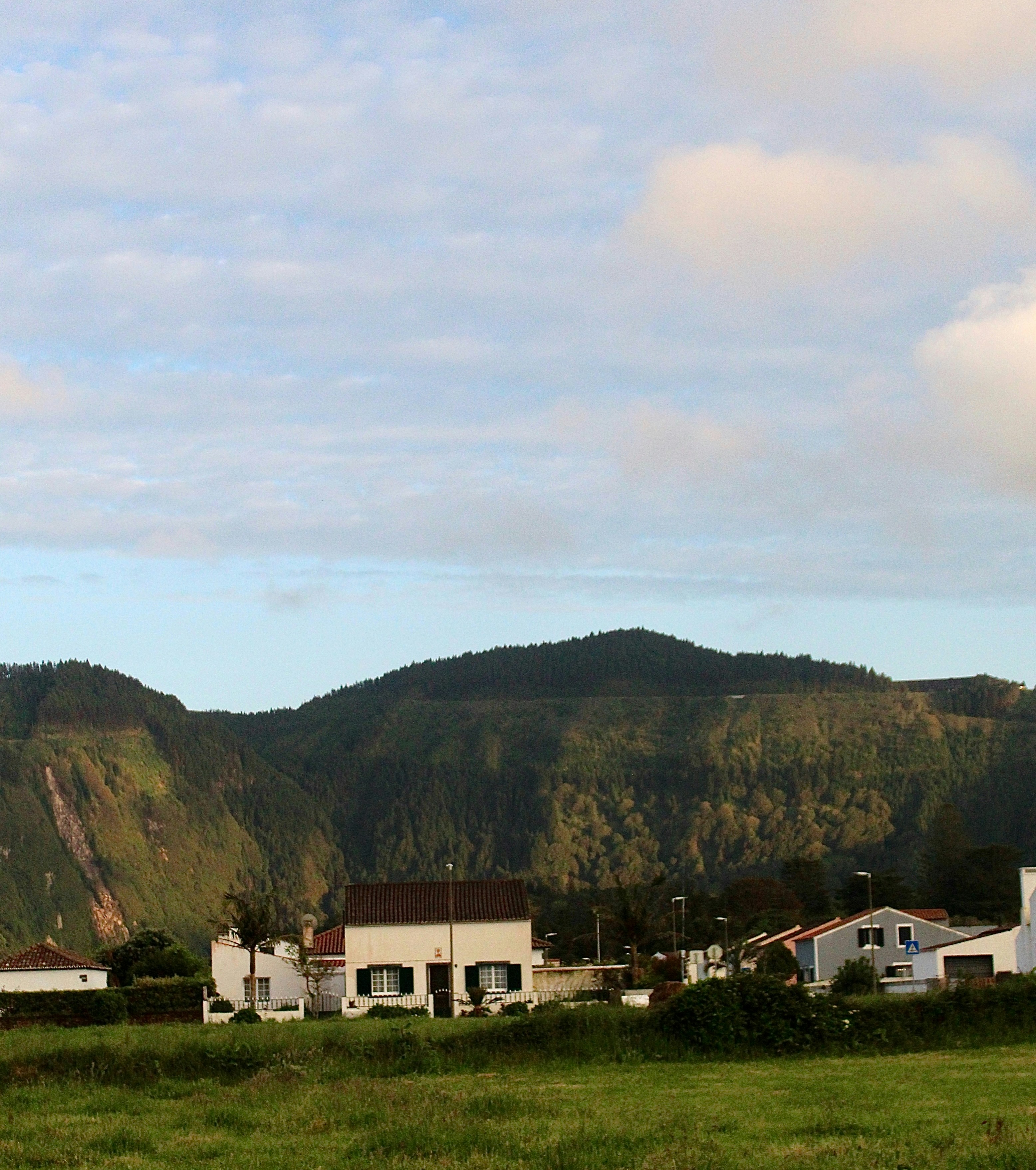 Quaint houses nestled against lush green hills under a partly cloudy sky. The scene captures the serene coexistence of human habitation and natural beauty.