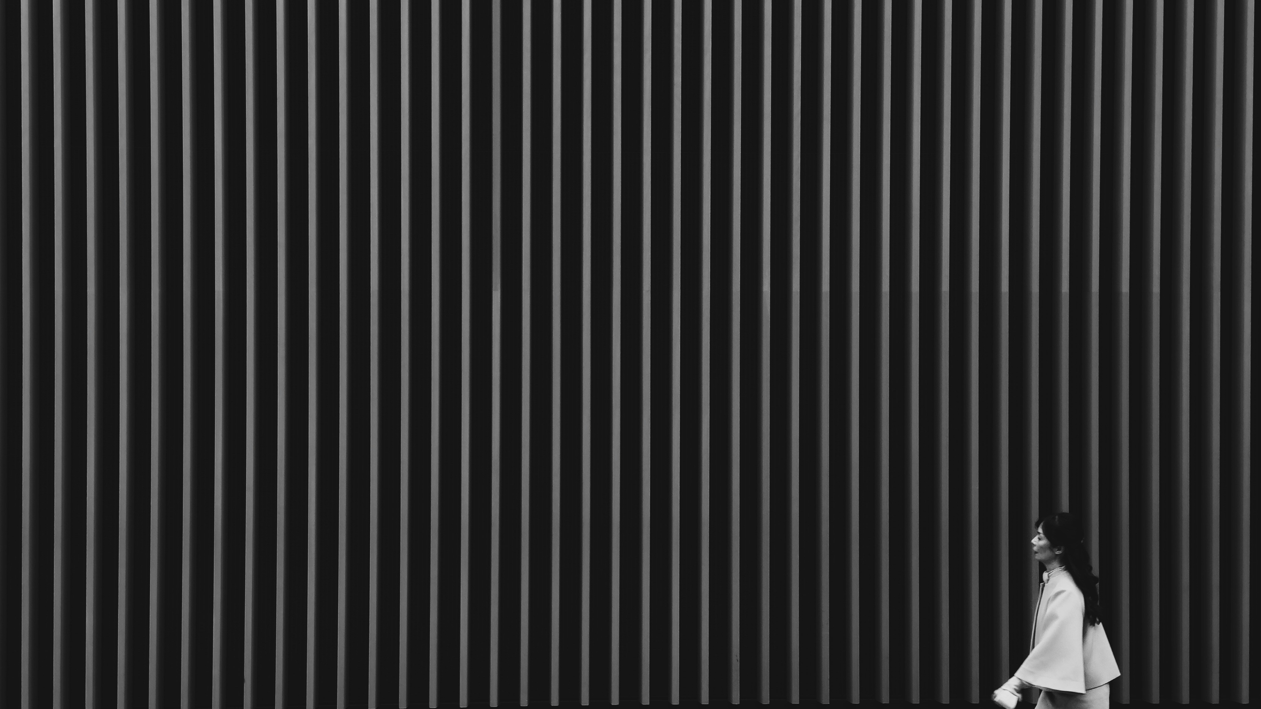 A woman walks past a series of vertical lines, creating a stark contrast in a minimalist setting. The monochrome palette emphasizes the interplay between light and shadow.