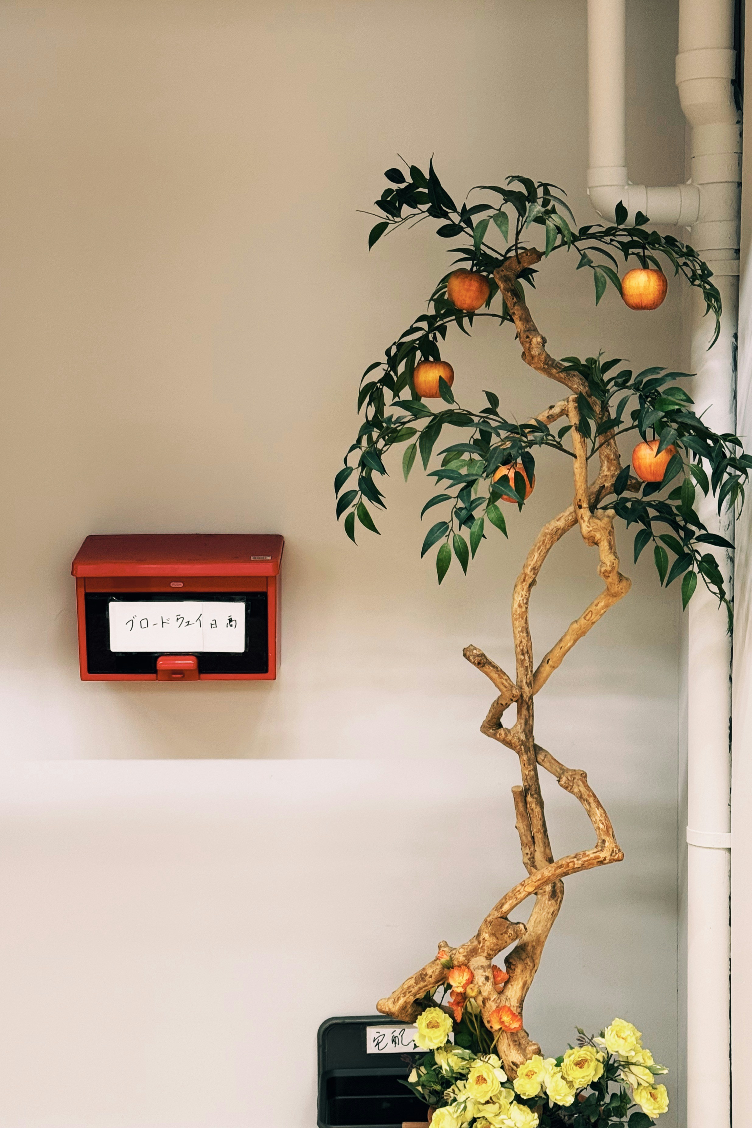 A creatively arranged faux tree adorned with artificial oranges stands beside a bright red mailbox labeled in Japanese. The scene combines elements of nature and urban life.