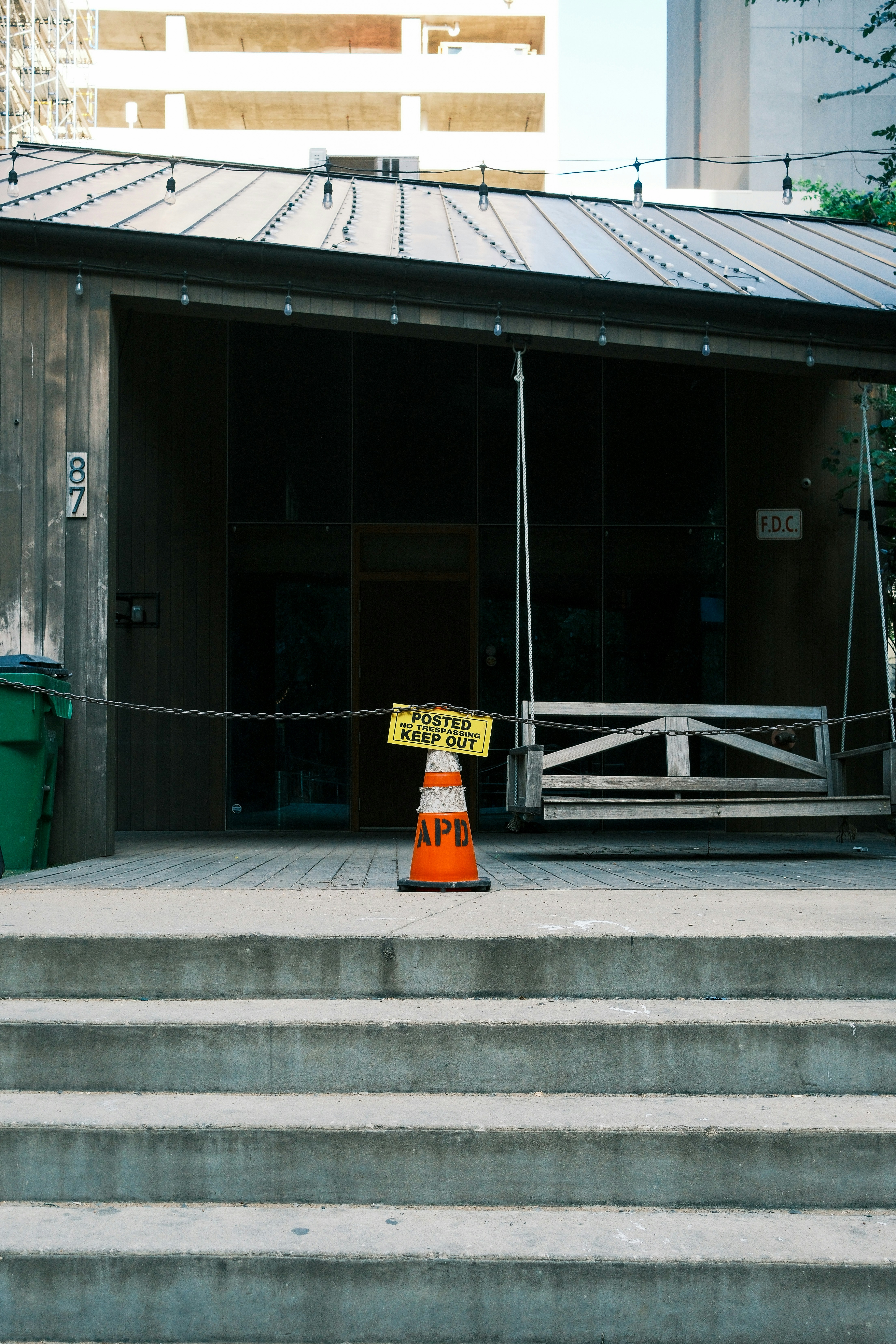 An empty swing and a cone marked APD outside a wooden structure in Austin. A scene of stillness, solitude, and subtle tension. | Building with caution tape, cone, and concrete stairs.