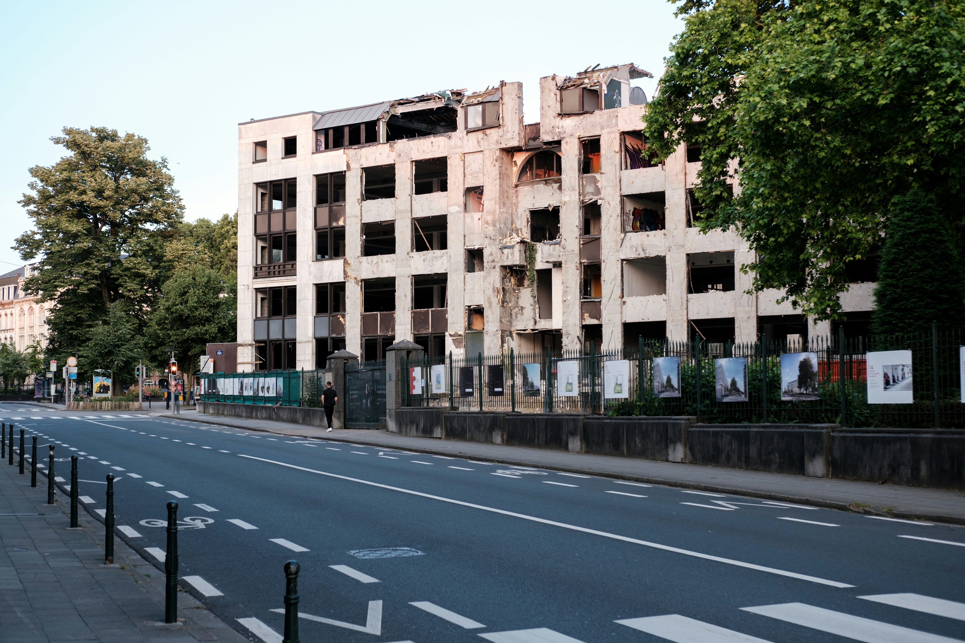 Decaying building with broken windows and peeling paint, surrounded by a quiet street and trees. Photographs displayed along the fence hint at its past.