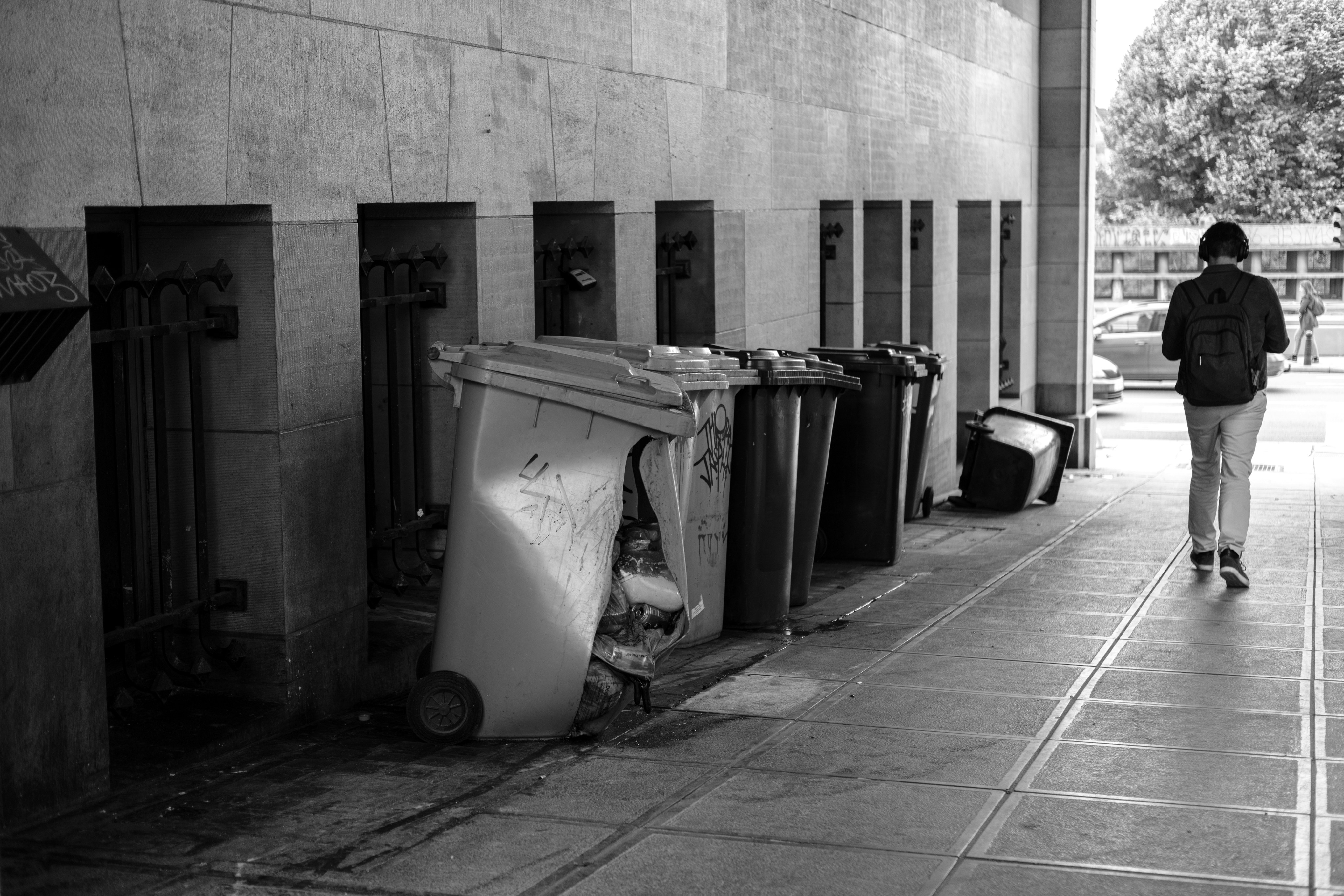 A solitary figure walks past a row of colorful trash bins in an urban alley, highlighting the contrast between human life and discarded remnants of daily activities.