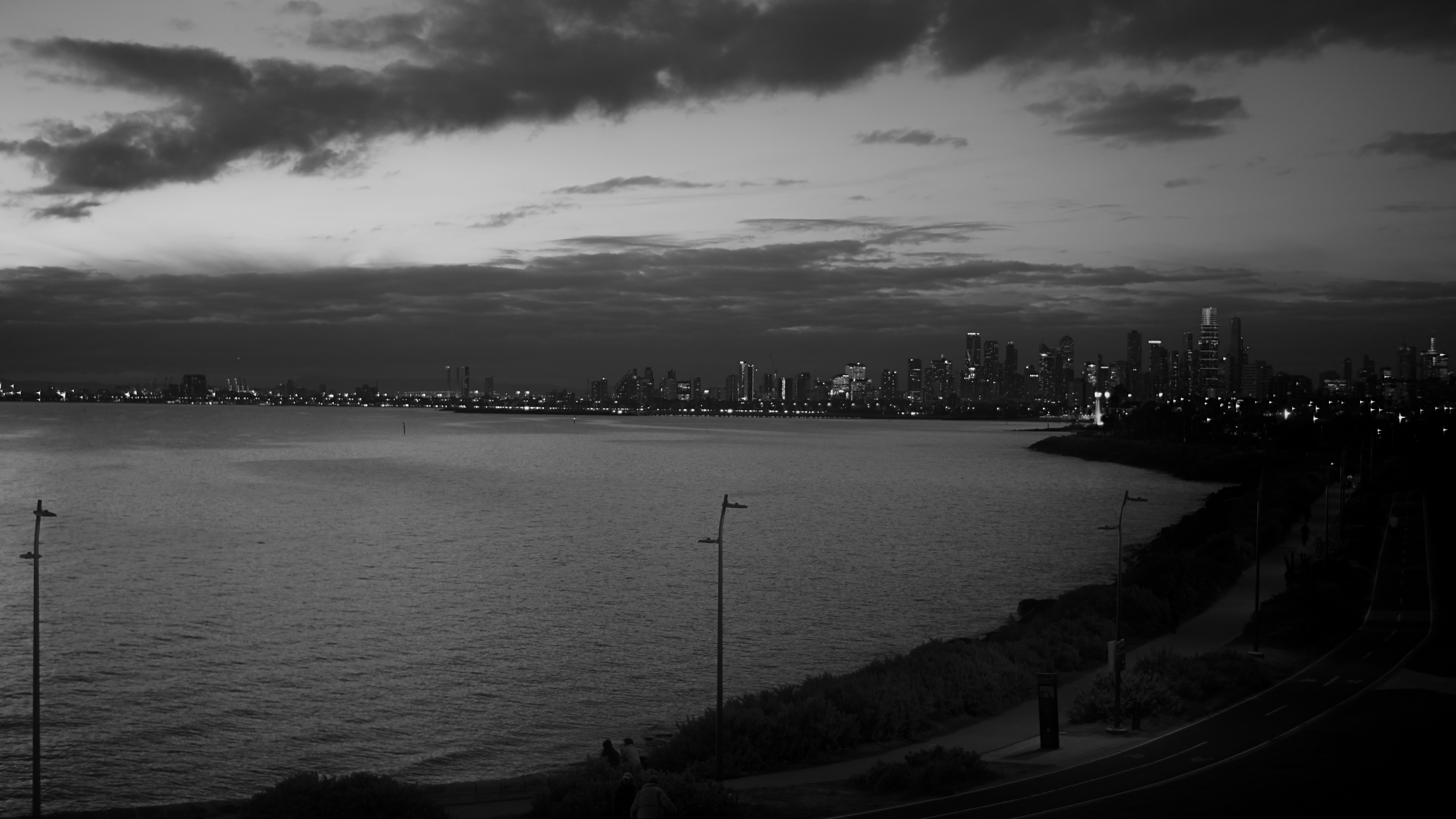 City skyline reflecting on water at twilight, with streetlights illuminating the shore. A serene blend of urban life and nature's tranquility.