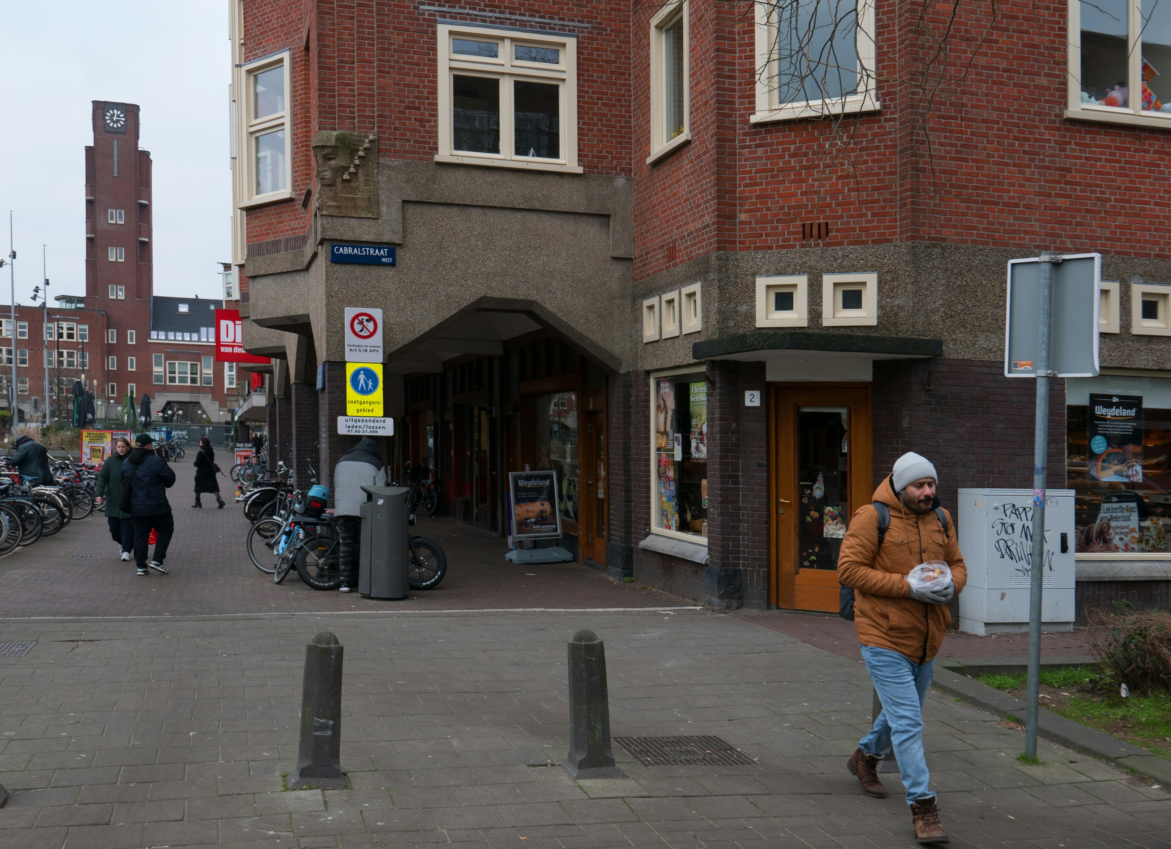 Free photo of a grey street view along the city square Mercatorplein in Amsterdam, in winter on a grey day in February. A man has shopped his food and walks home over the wide sidewalk with brick pavement. Street photography, free image CC0 & high resolutions by Fons Heijnsbroek, The Netherlands Dutch / Nederlands: 2025 - gratis download foto van het Mercatoplein in Amsterdam op een grijze dag in februari. Een man loopt over de brede stoep met achter hem de bakstenen lage passage. Amsterdamse School!! - straatfotografie - foto in hoge resolutie CC0, Fons Heijnsbroek, Nederland. | A street corner with brick buildings and people.