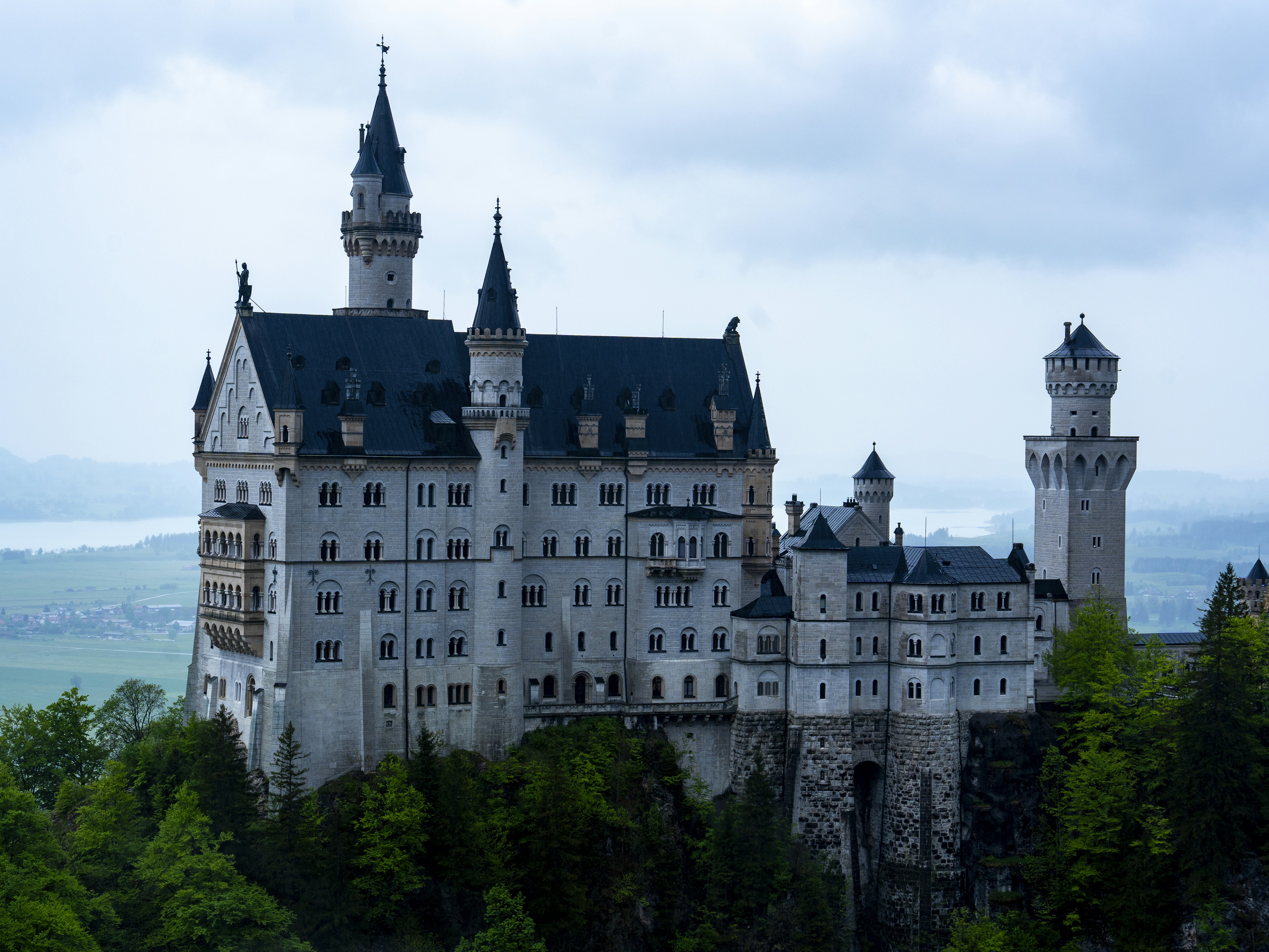 Neuschwanstein Castle perched on a hillside, surrounded by lush greenery and distant mountains under a cloudy sky.