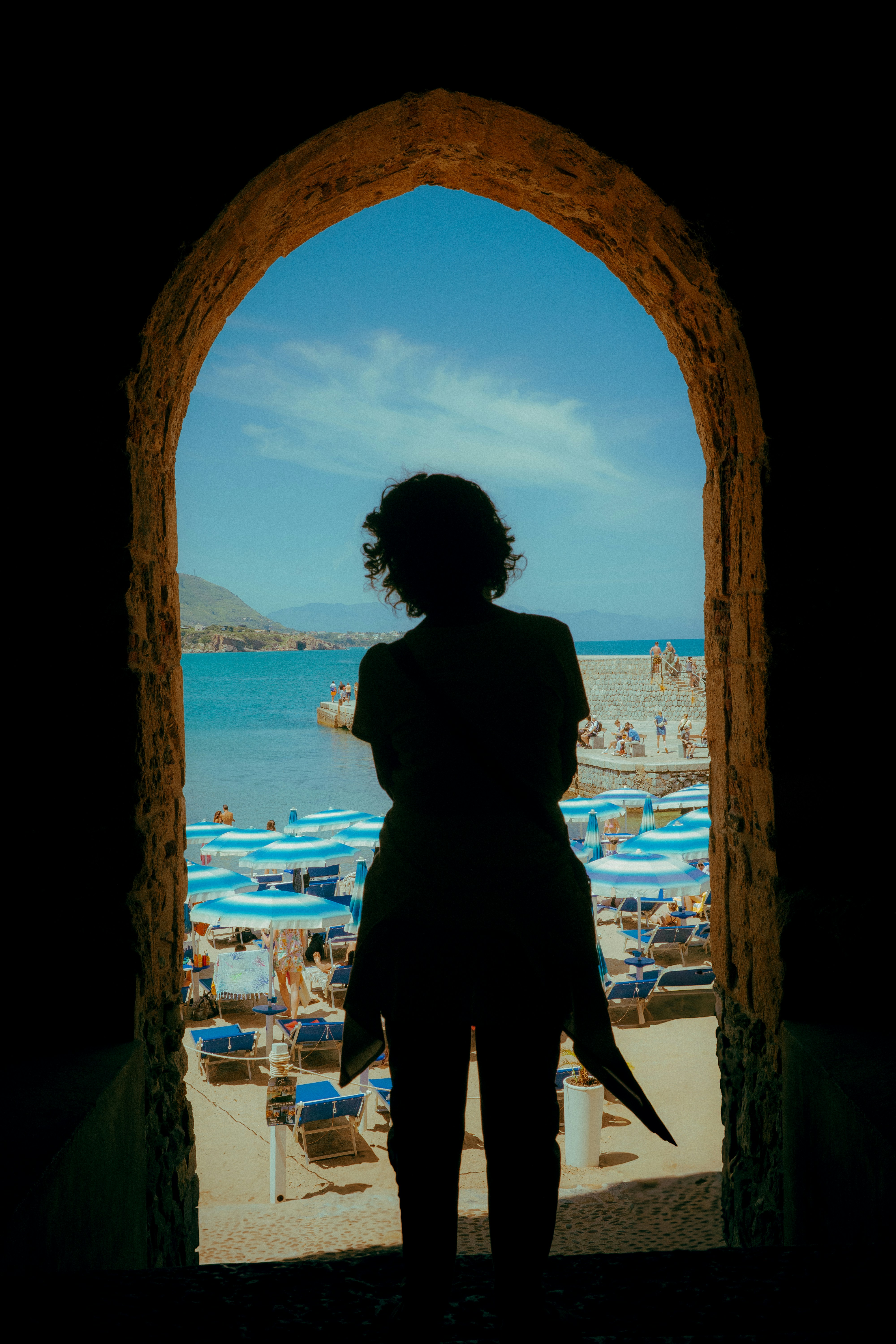 Silhouette in a Stone Arch Framing a Sunlit Beach with Azure Umbrellas