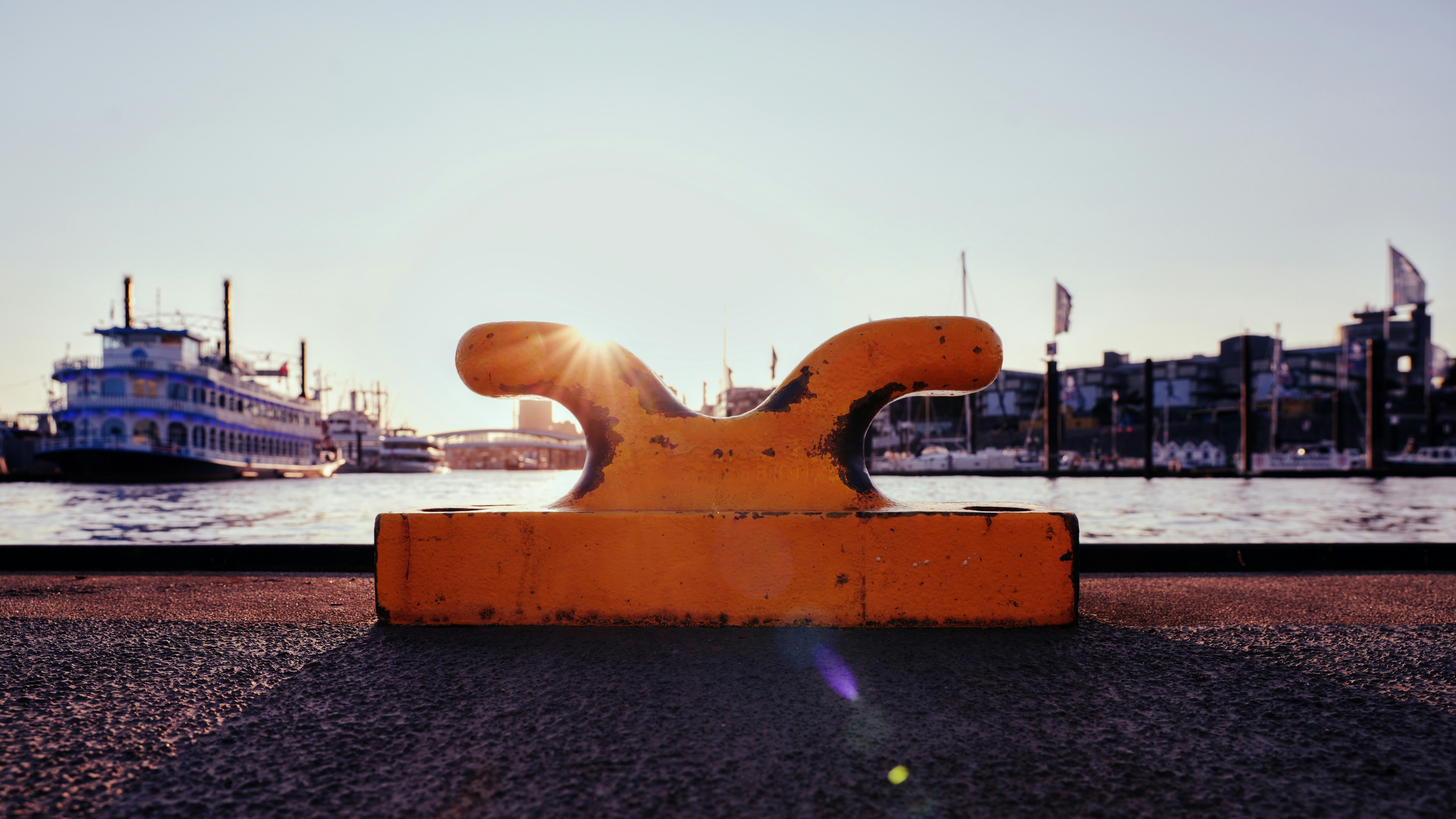 A vibrant yellow cleat stands in the foreground, framing a bustling harbor scene with boats and buildings in the background. The sun sets, casting a warm glow over the water.