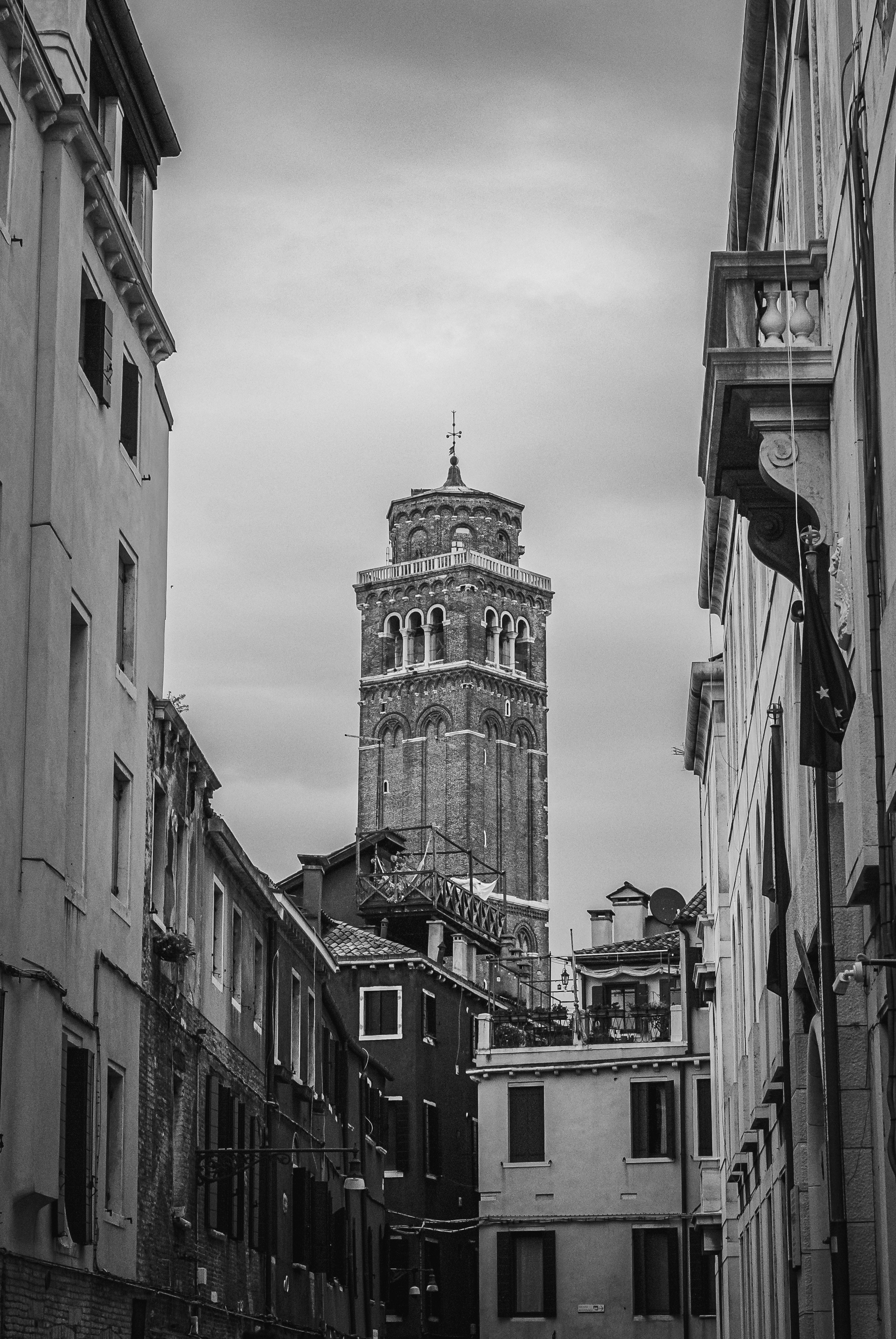 A historic bell tower rises above narrow Venetian streets, framed by aged buildings under a moody sky.