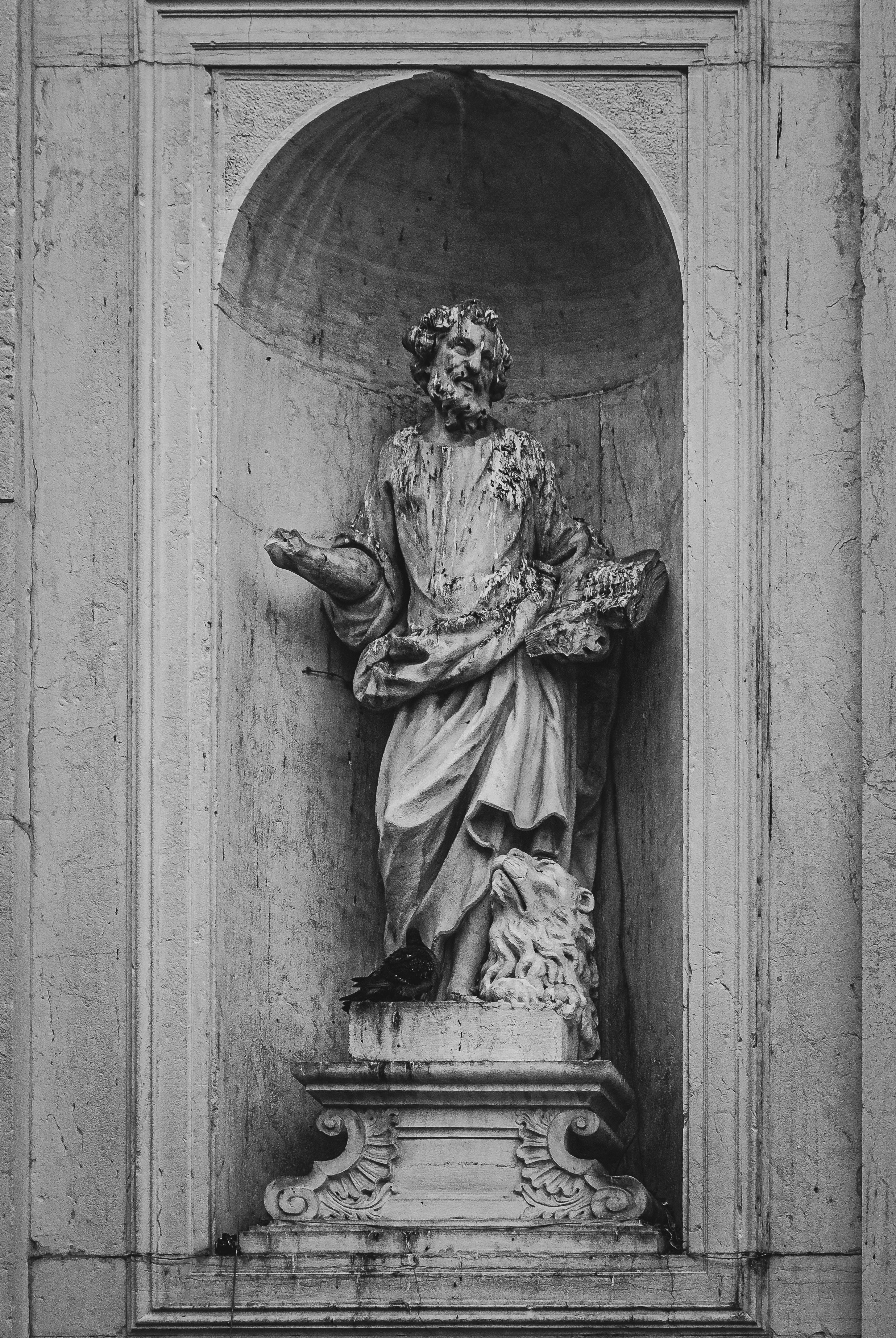 A weathered statue of a figure with outstretched arms, adorned with intricate details, set within an ornate niche. A small bird perches at its base.