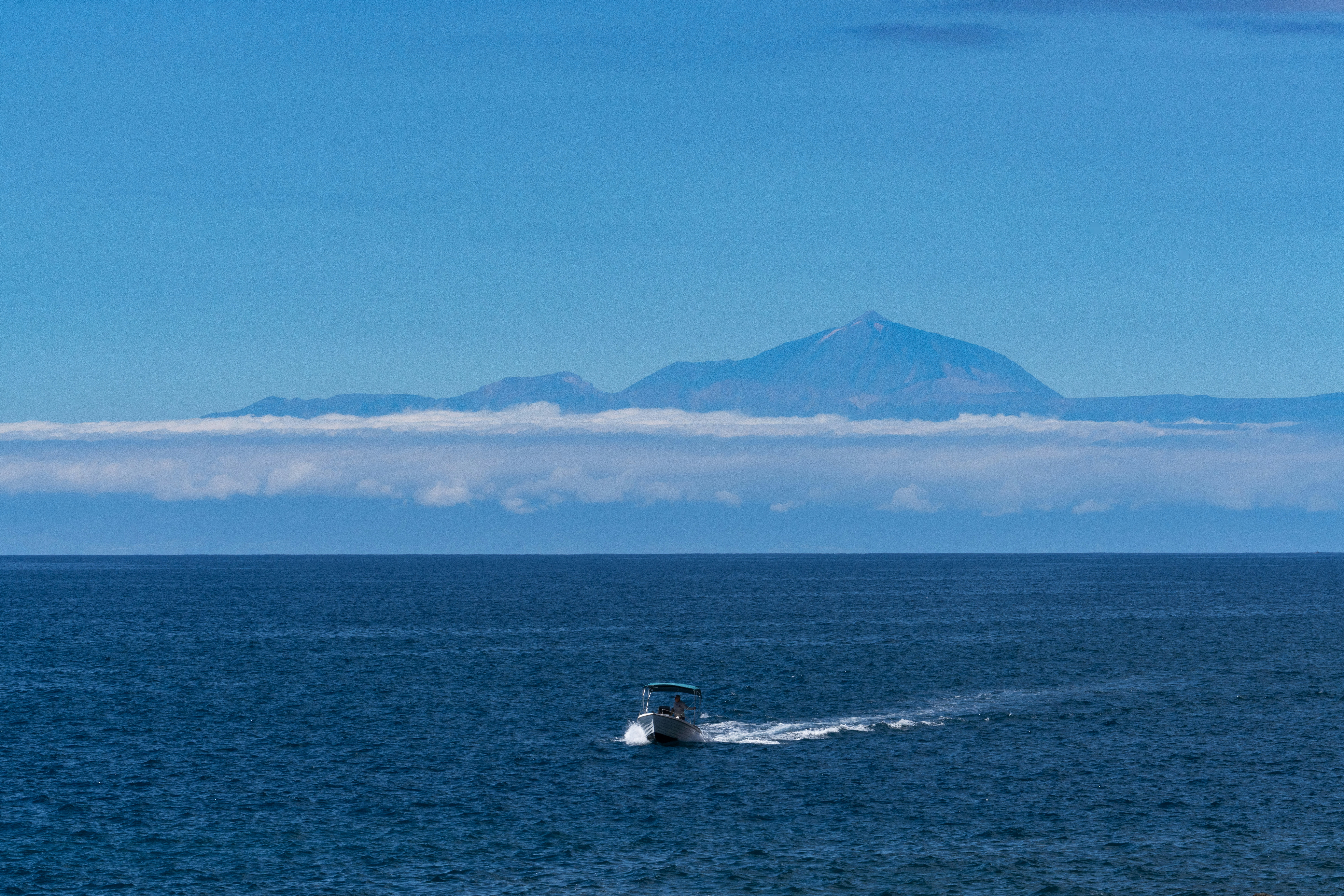 A small boat navigates the tranquil ocean waters, with a distant mountain range shrouded in clouds on the horizon.