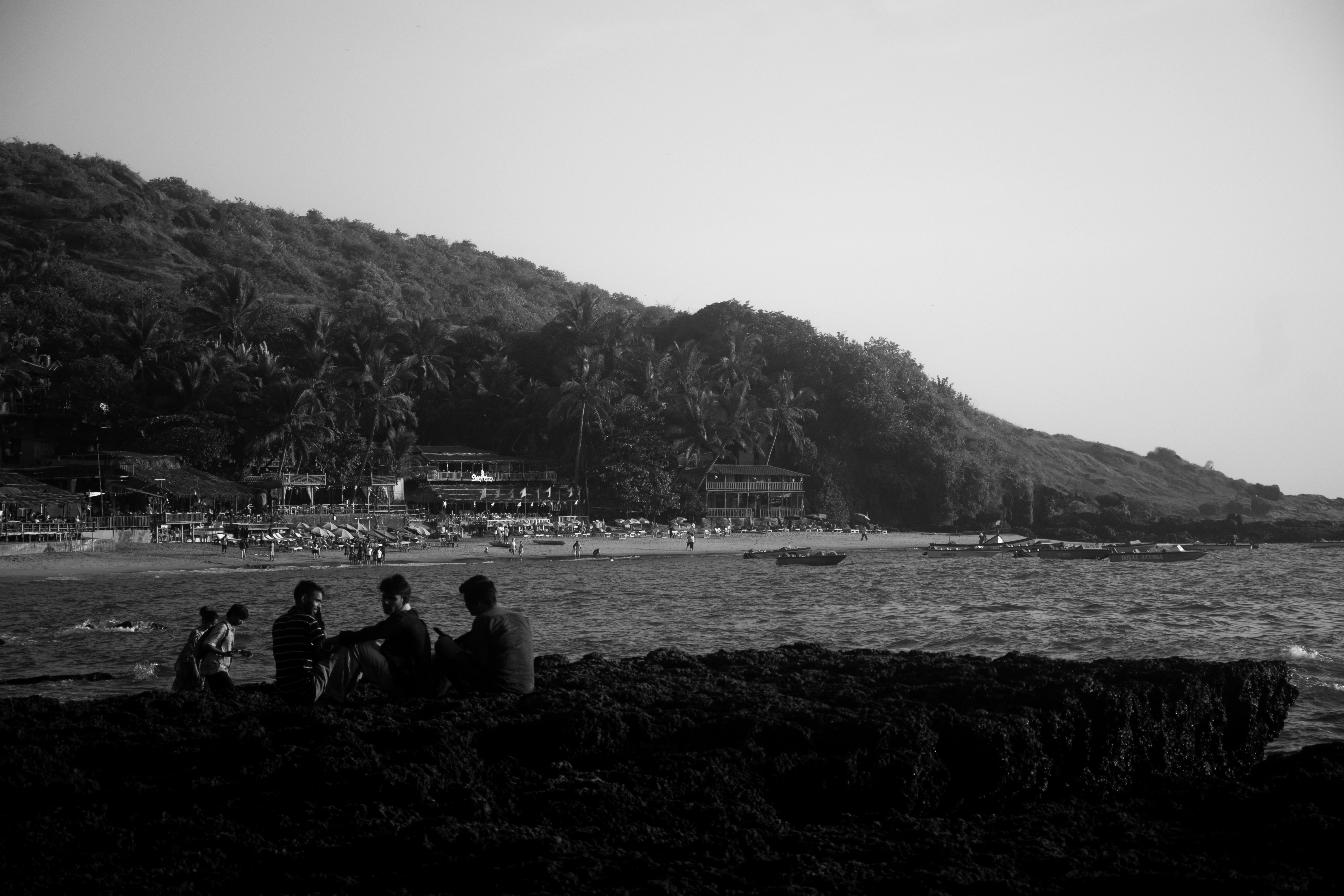 Group of friends gathered on rocky beach, engaging in conversation as the sun sets behind a lush hillside. 