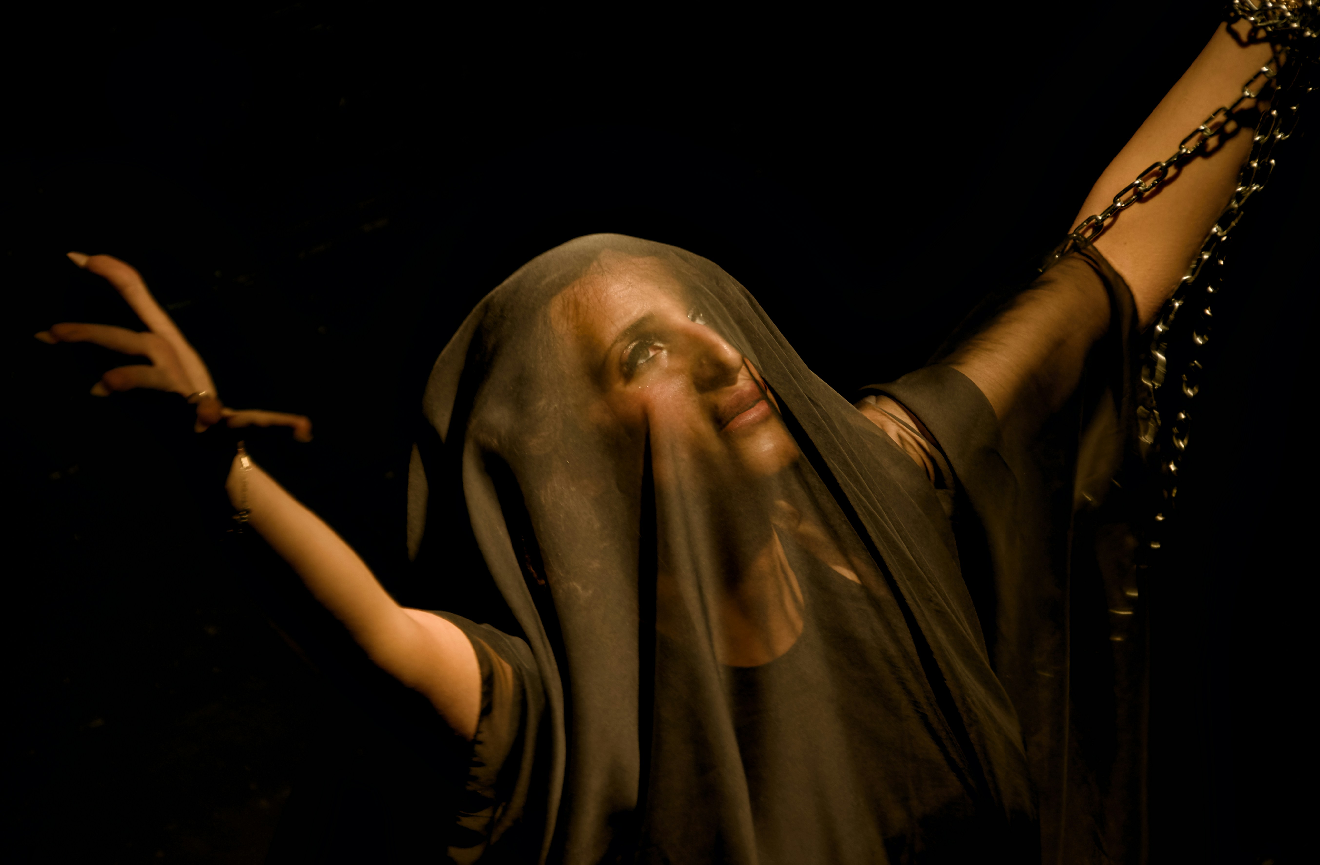 A figure draped in a sheer veil extends their arms, evoking a sense of liberation amidst darkness. The interplay of light and shadow enhances the dramatic atmosphere.
