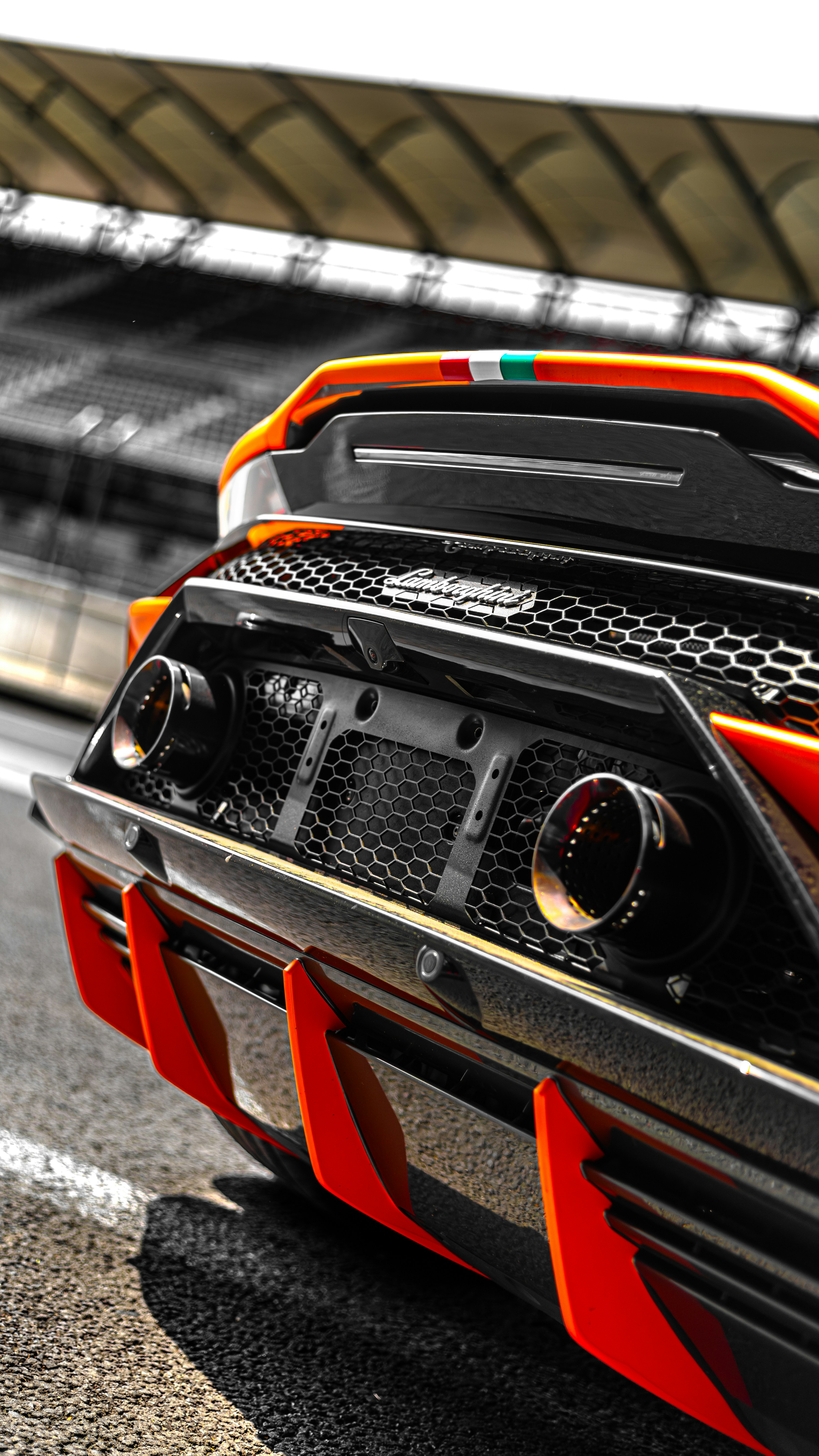 Close-up view of a Lamborghini's rear detailing, showcasing its aerodynamic design and dual exhausts. The background features a blurred racetrack setting.