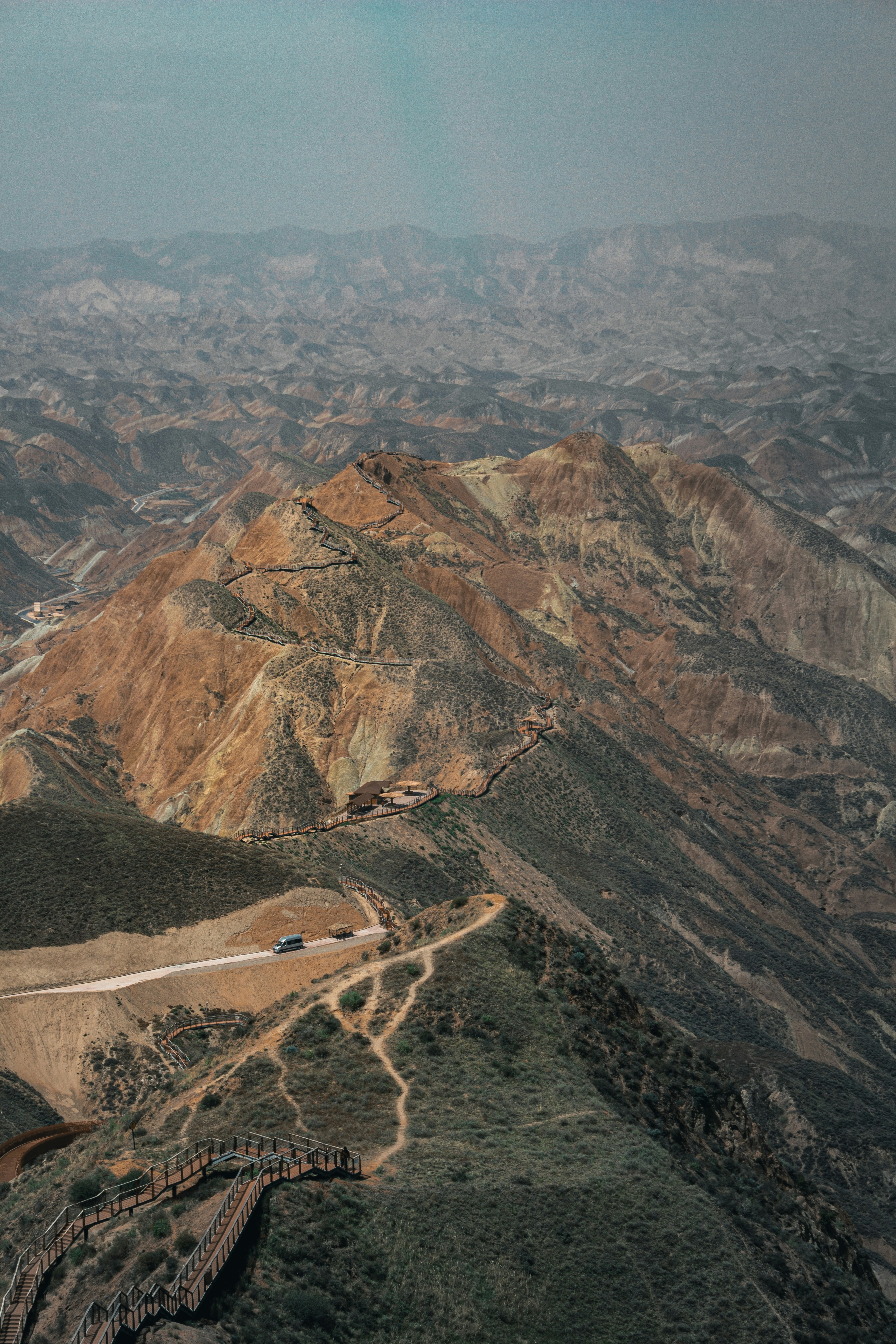 Aerial view of undulating hills with vibrant earth tones, featuring winding paths and a distant vehicle on a dirt road.