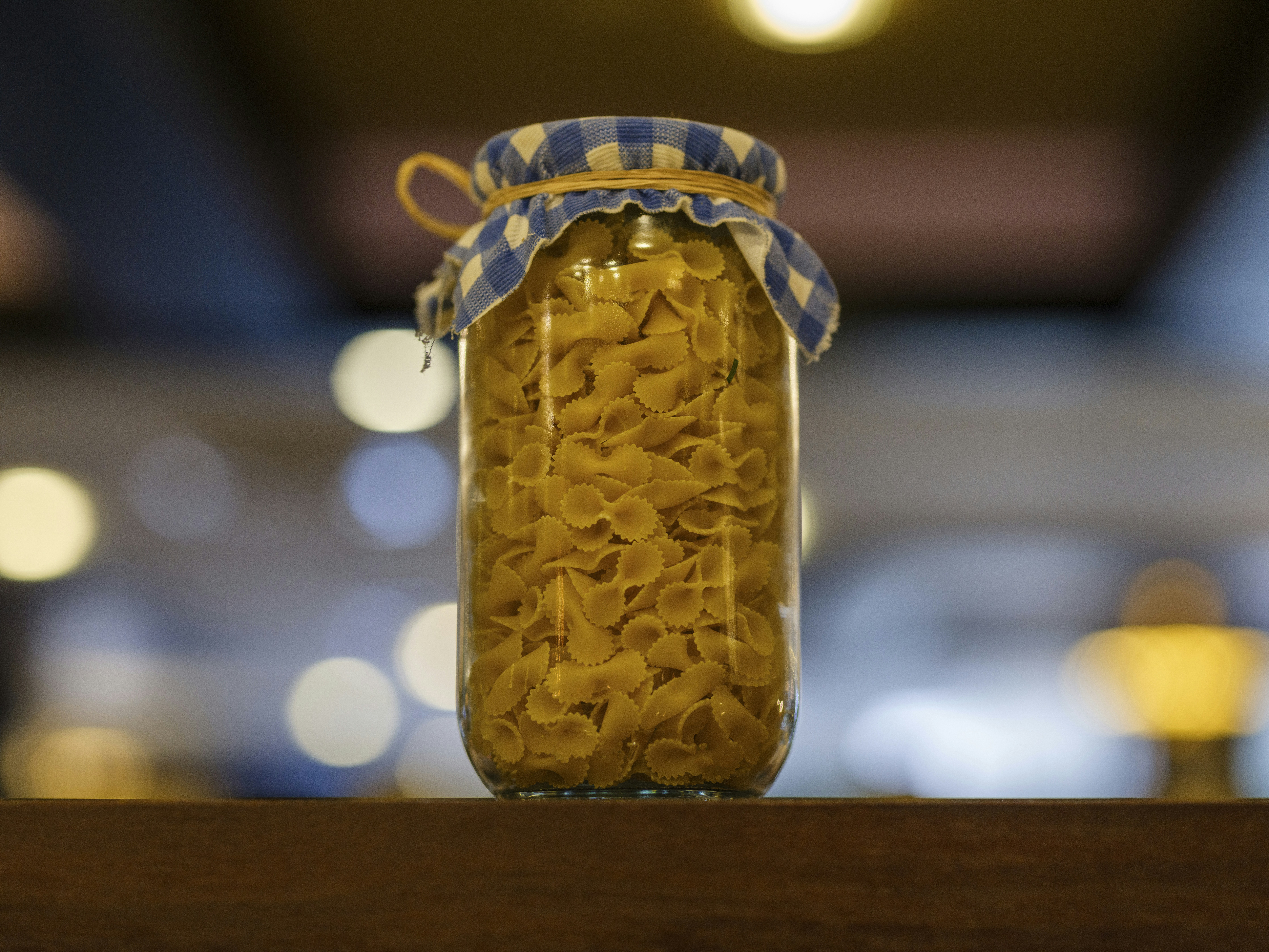 Glass jar filled with bow-tie pasta, adorned with a blue checkered lid, set against a softly blurred background. 