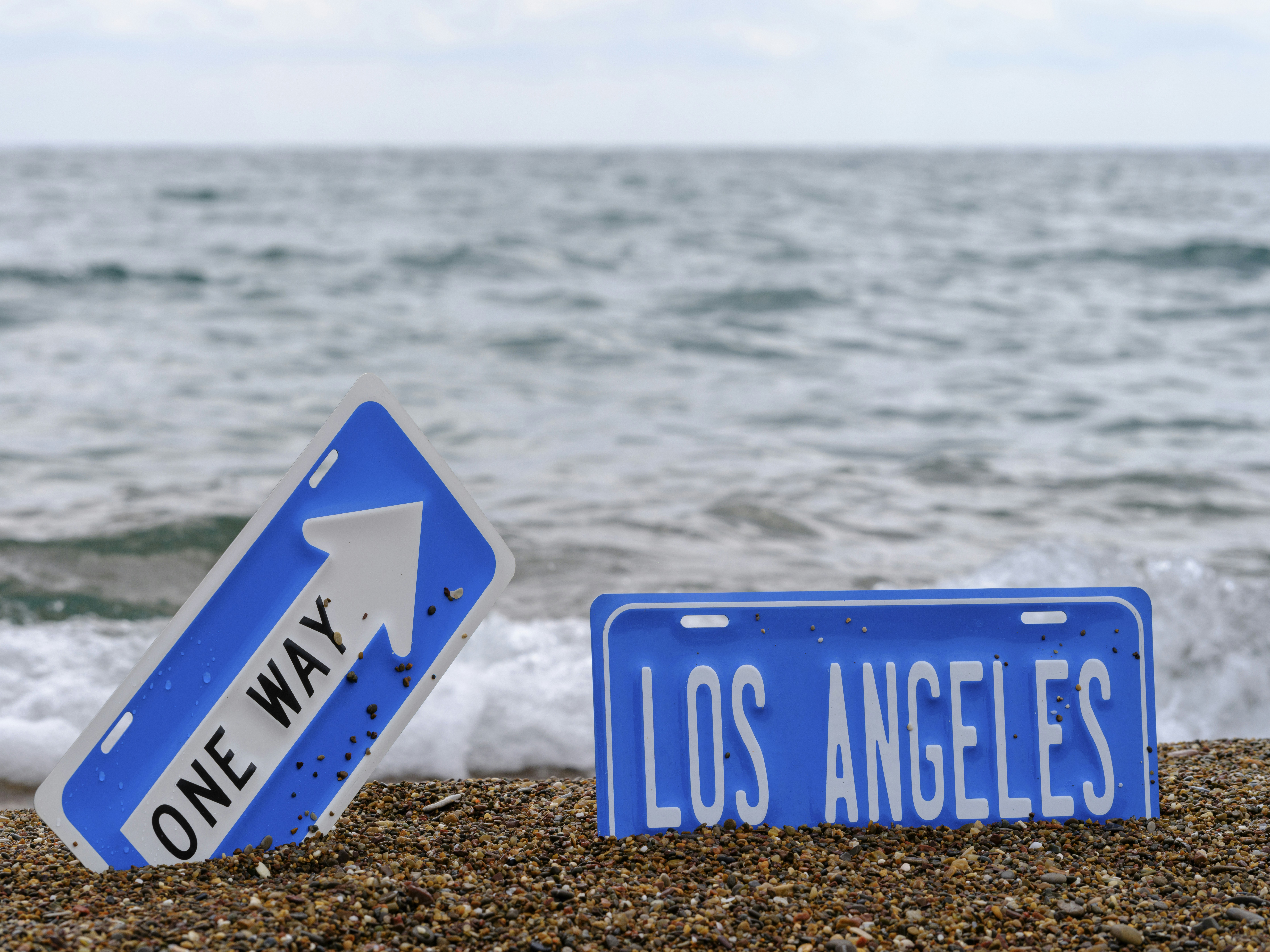 Blue Signposts by the Pacific: Tilted One Way and Los Angeles on a Pebbled Beach