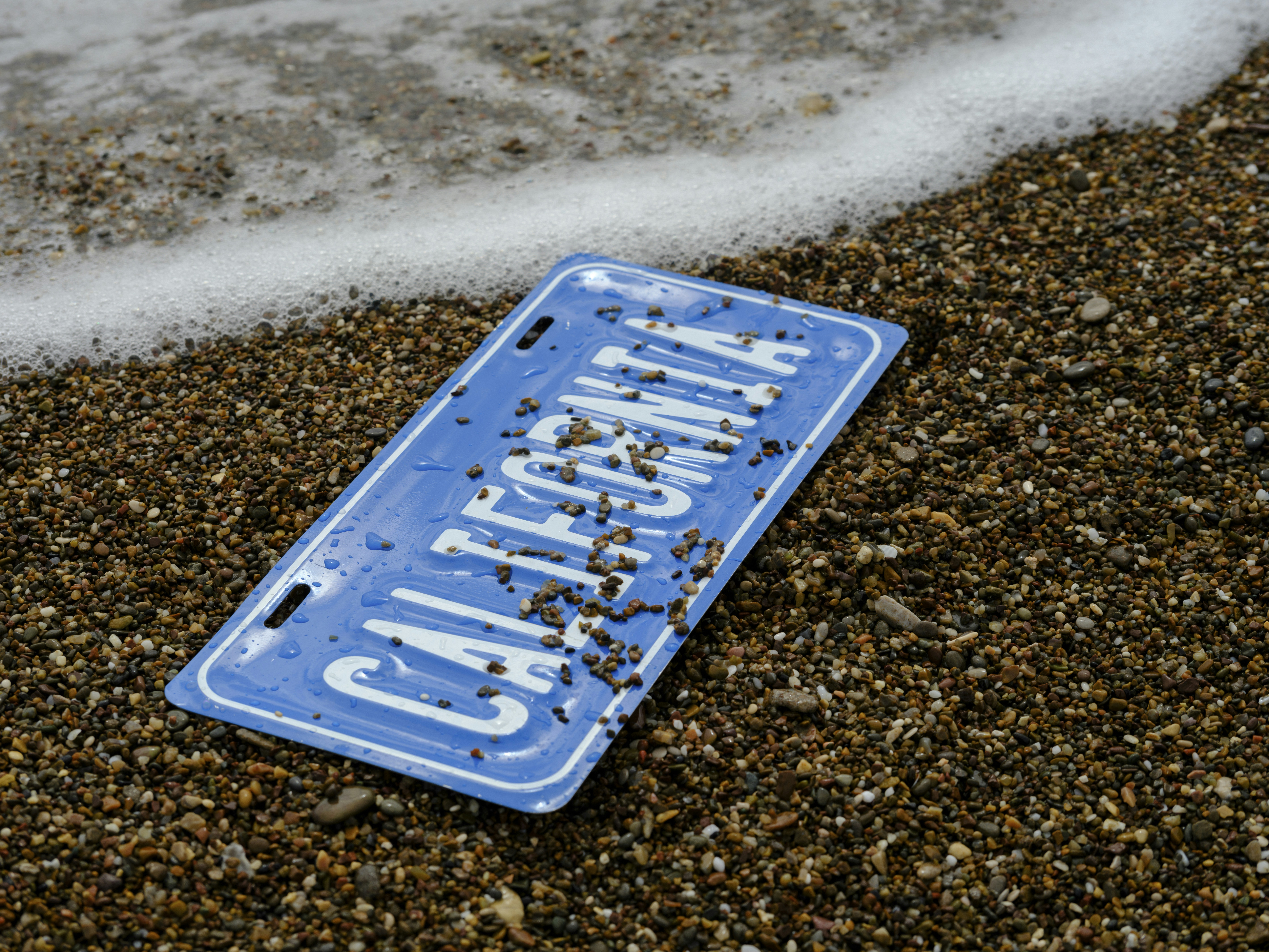 A weathered California license plate partially buried in pebbles, with gentle waves lapping at its edges.