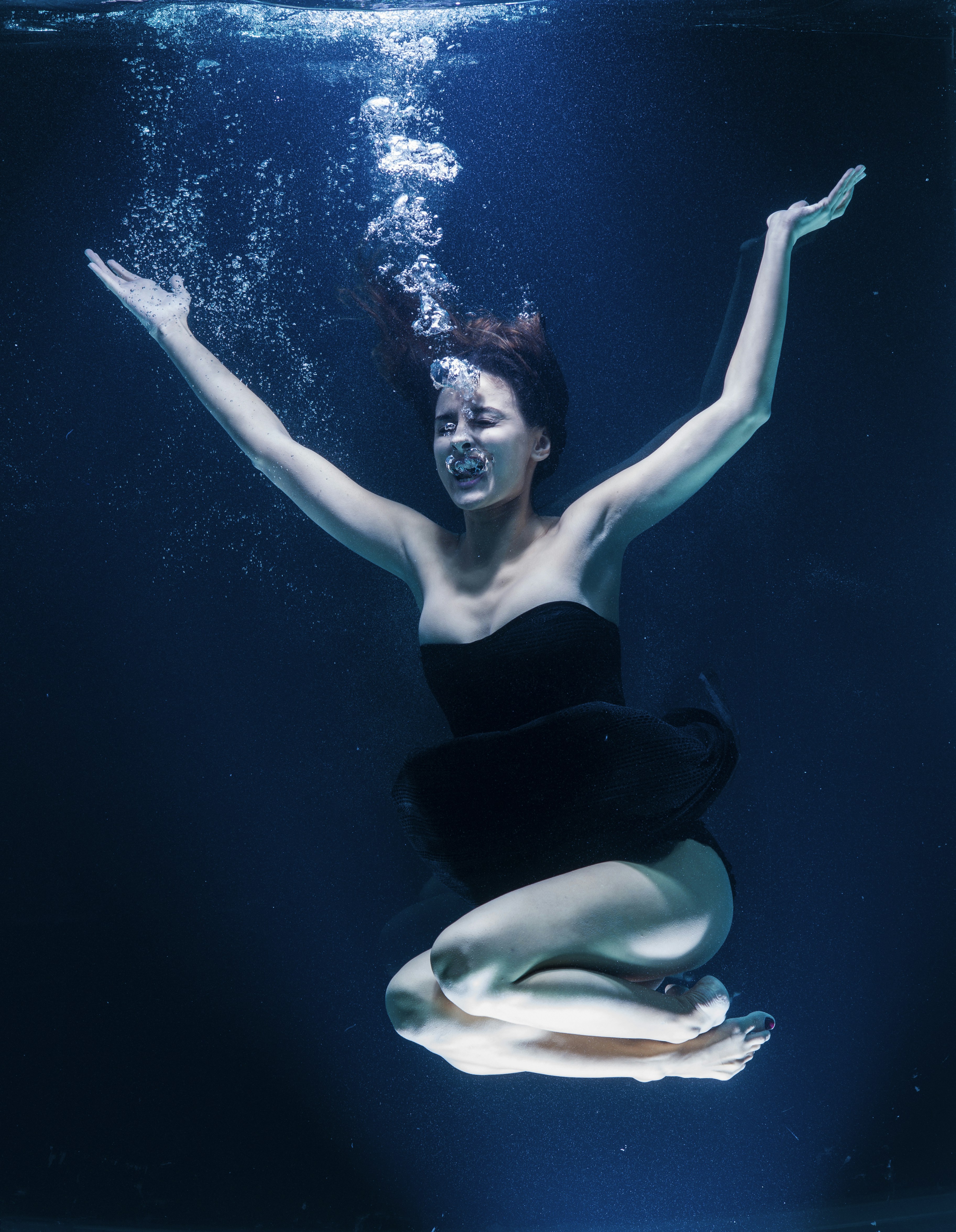 A woman in a black dress gracefully floats underwater, surrounded by bubbles and dark blue water, embodying a moment of serene freedom.