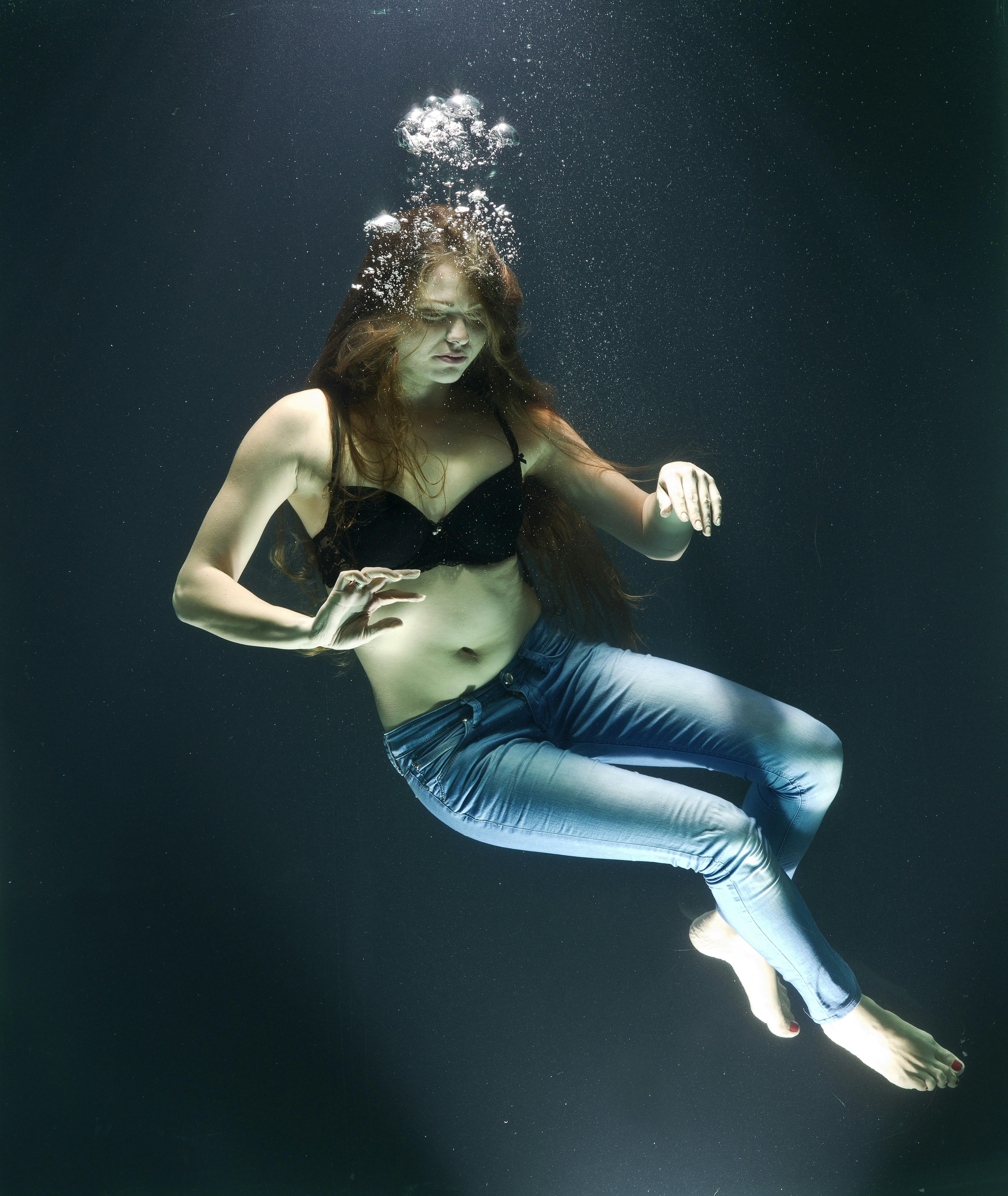A young woman gracefully floats underwater, embodying tranquility in a dark aquatic environment. Her flowing hair and casual attire contrast with the surrounding water bubbles.