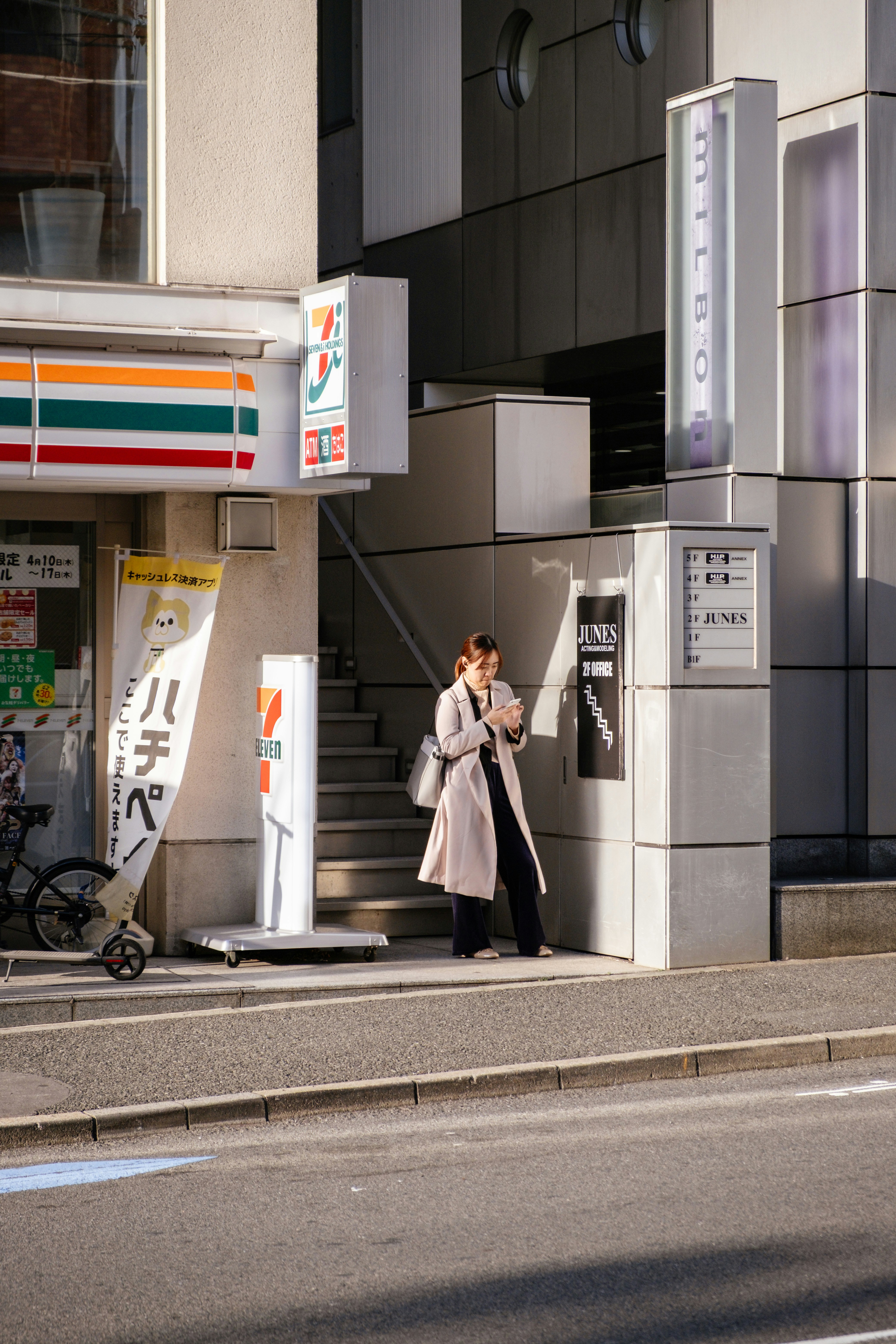 A woman in a long coat stands on a city sidewalk, absorbed in her phone, with a 7-Eleven store and modern architecture in the background.