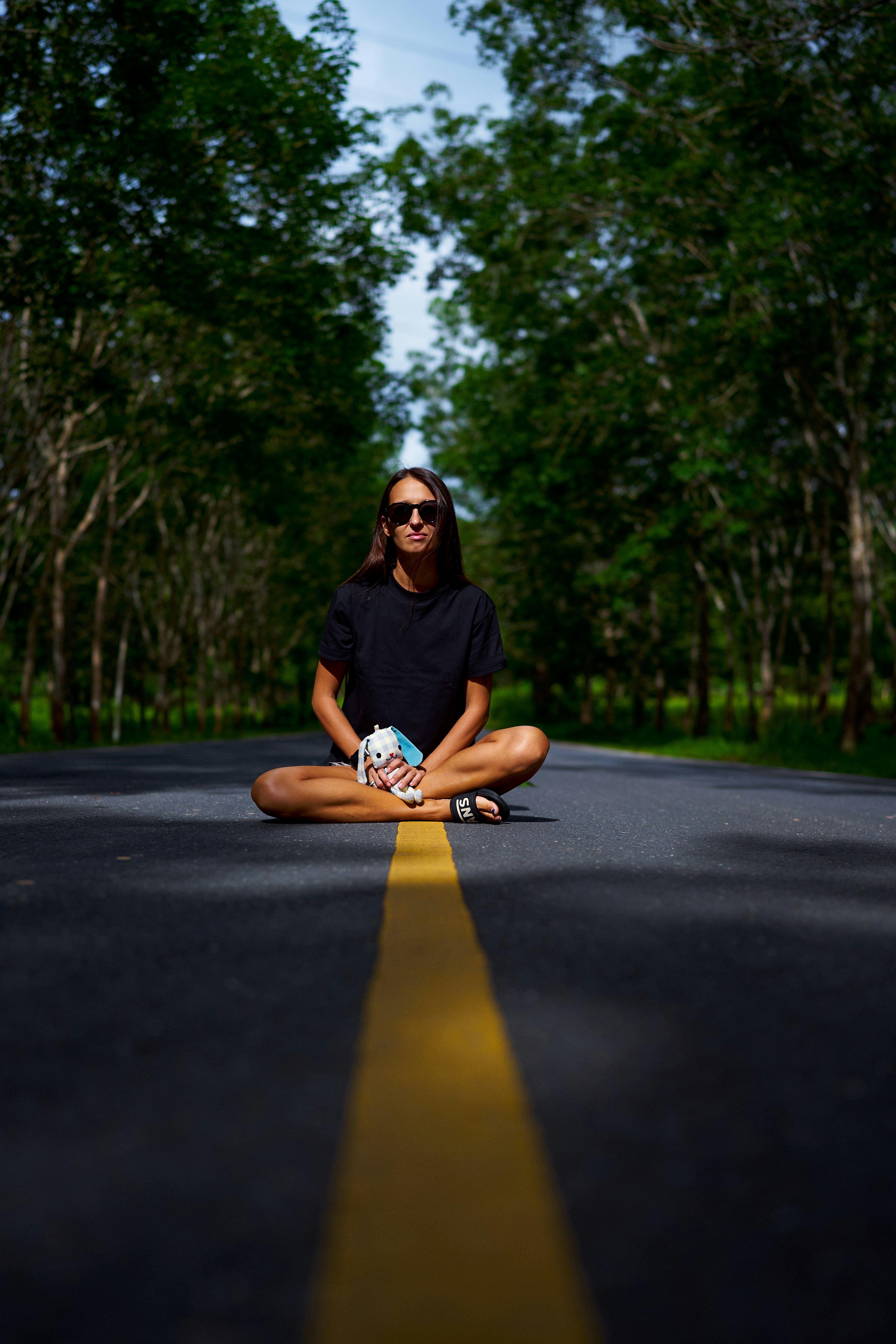Young woman seated cross-legged on a quiet road, holding a small dog, surrounded by lush greenery.