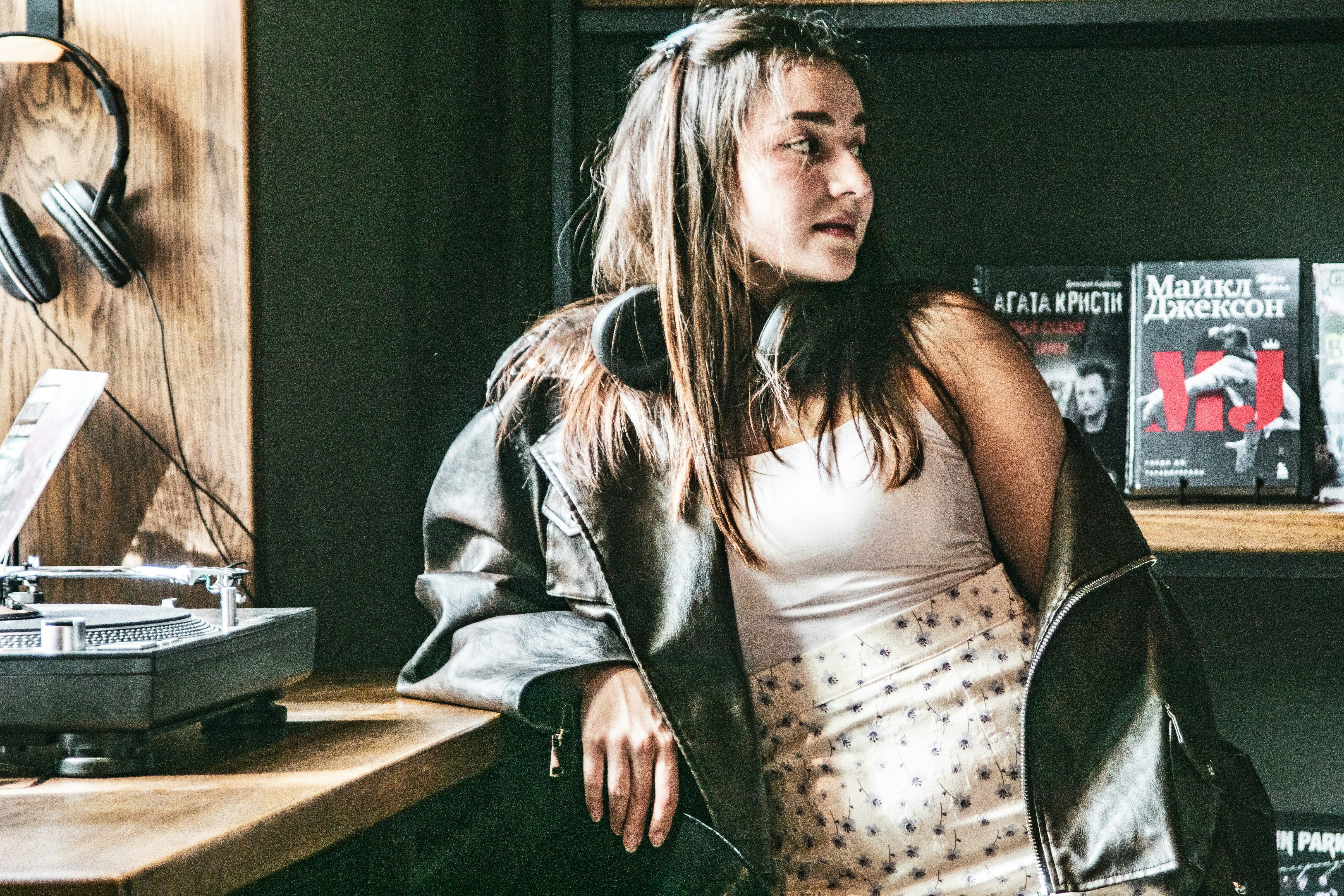 A girl in a leather jacket and a white tank top is standing in front of the interior of a record store.