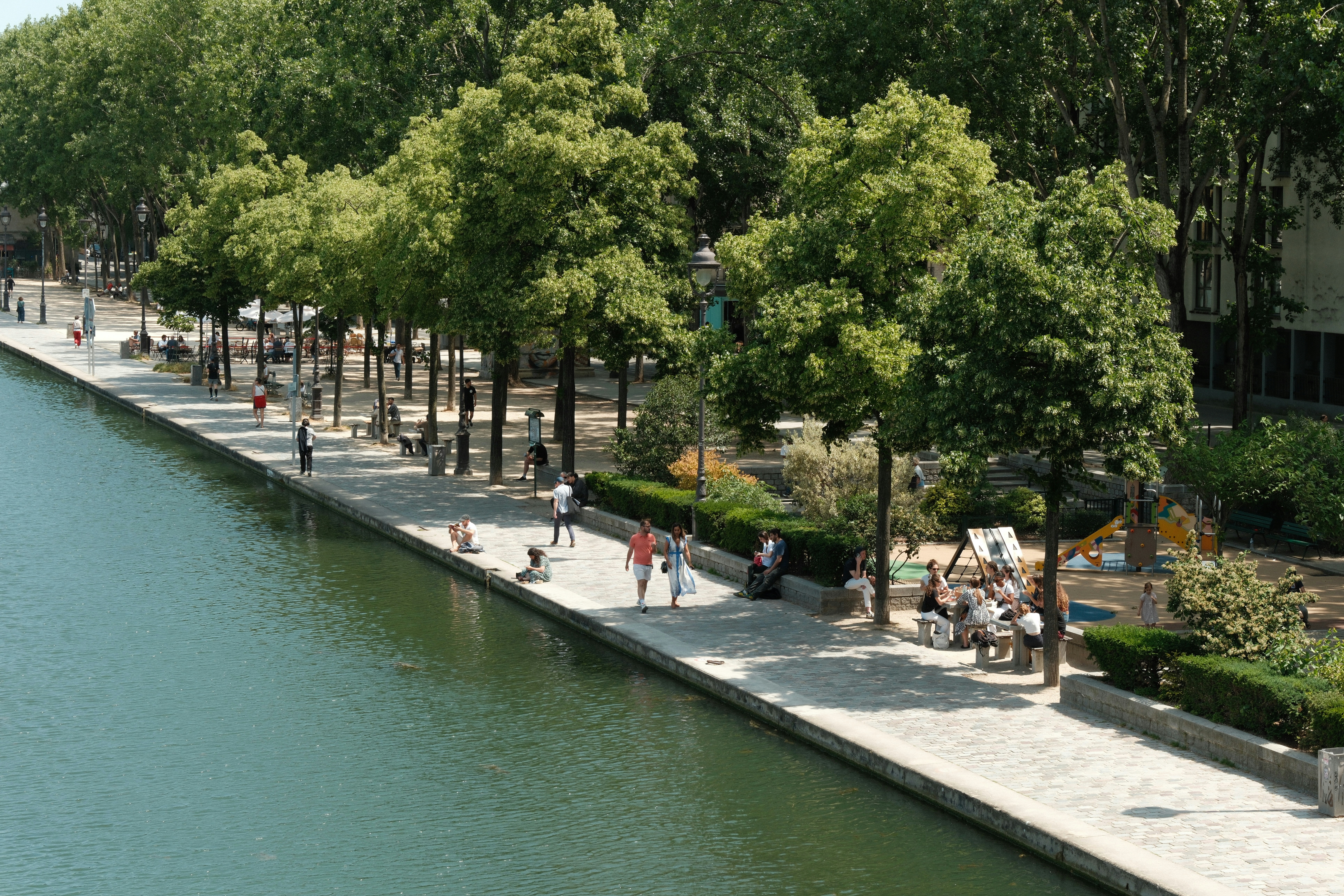 wide shot of the side of a canal in Paris 19th, with groups of people walking along the water and the trees