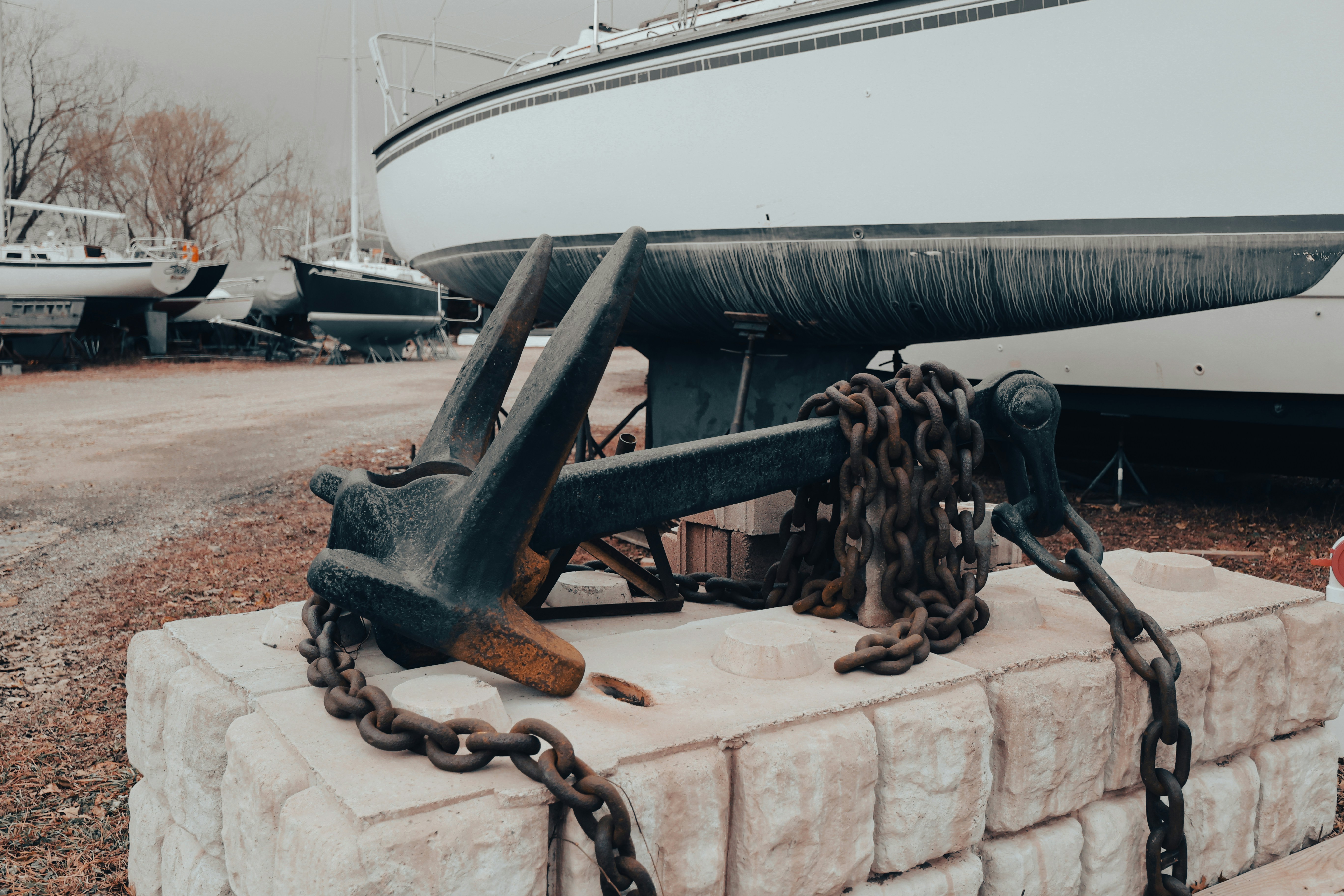 A large, weathered anchor rests on a stone block near docked sailboats, its heavy chain coiled in rusted loops. | Anchor with chains, in front of a boat.