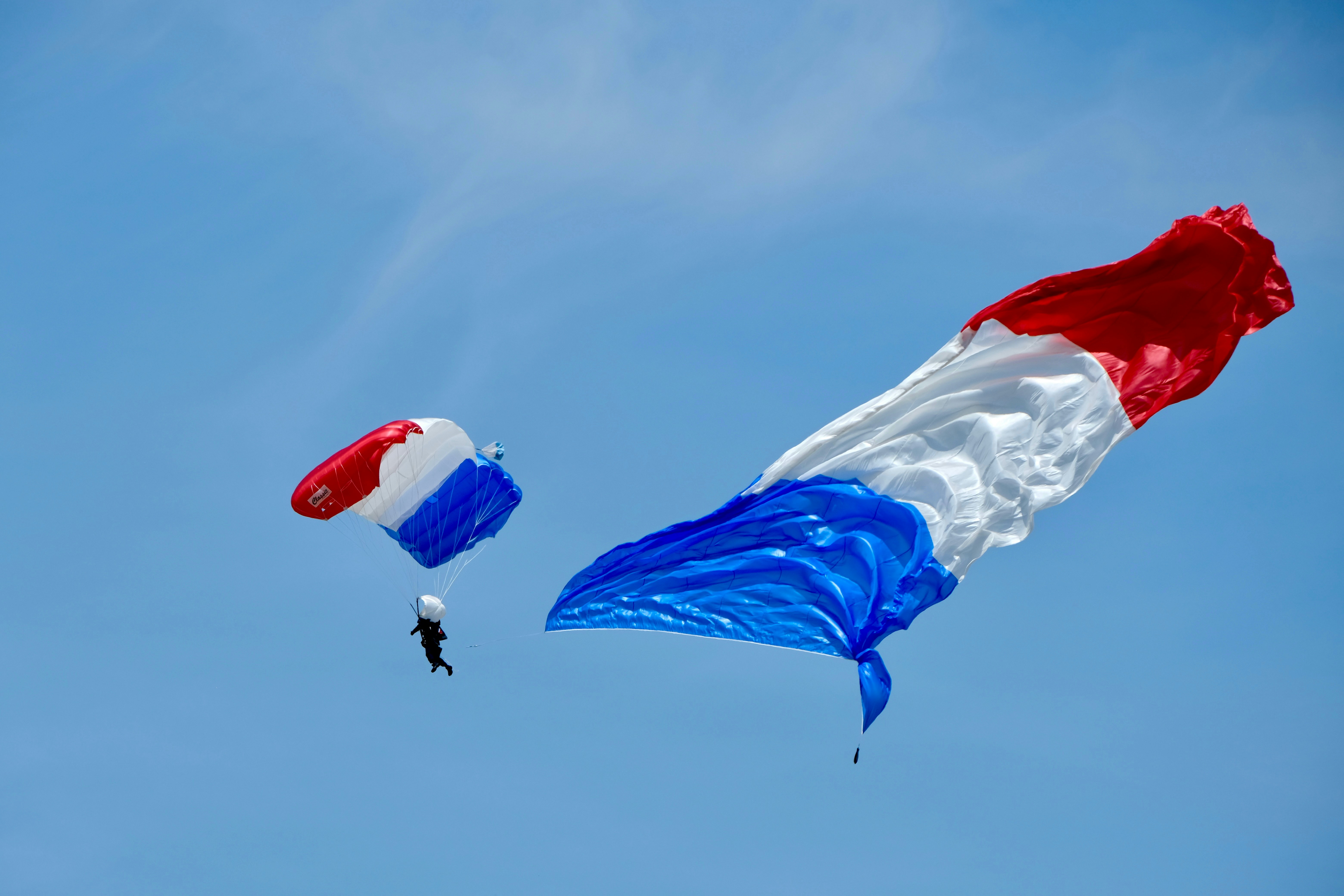 A parachutist descends gracefully beneath a vibrant tricolor parachute, with a larger flag billowing above against a clear blue sky.