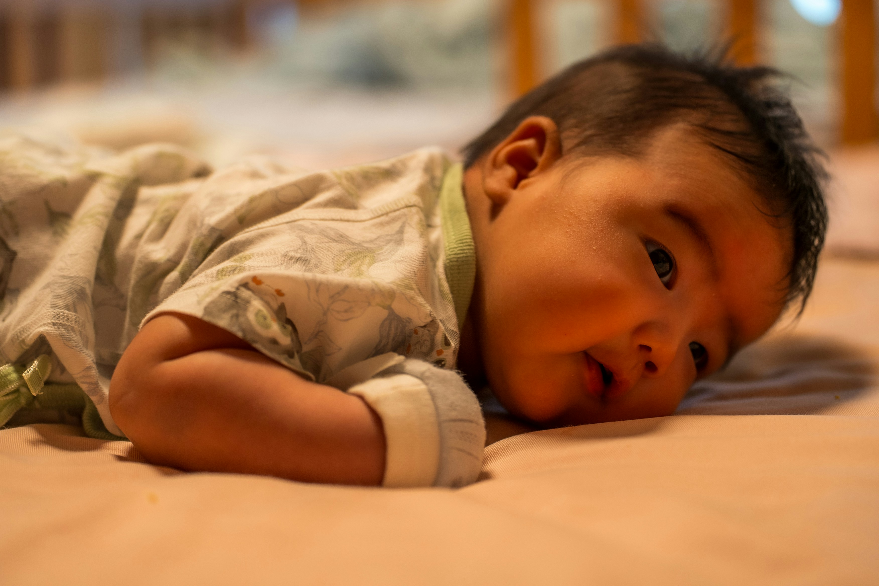 A baby lying on a soft surface, displaying a curious expression while propped on their arms. The gentle light casts a warm glow, enhancing the serene atmosphere.